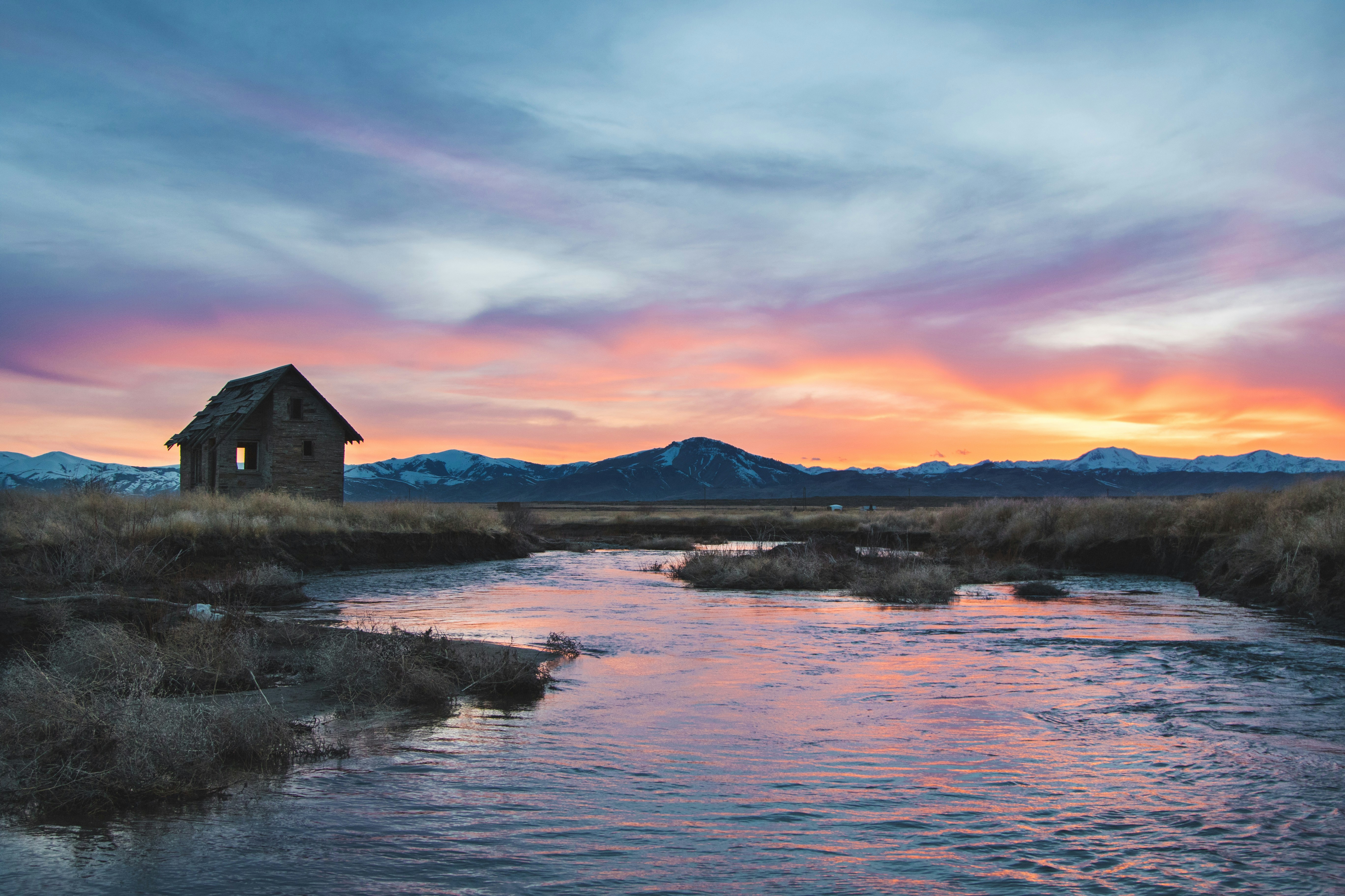 Abandoned wooden cabin beside a gently flowing river under a vibrant sunset sky. The scene captures the essence of solitude in nature.