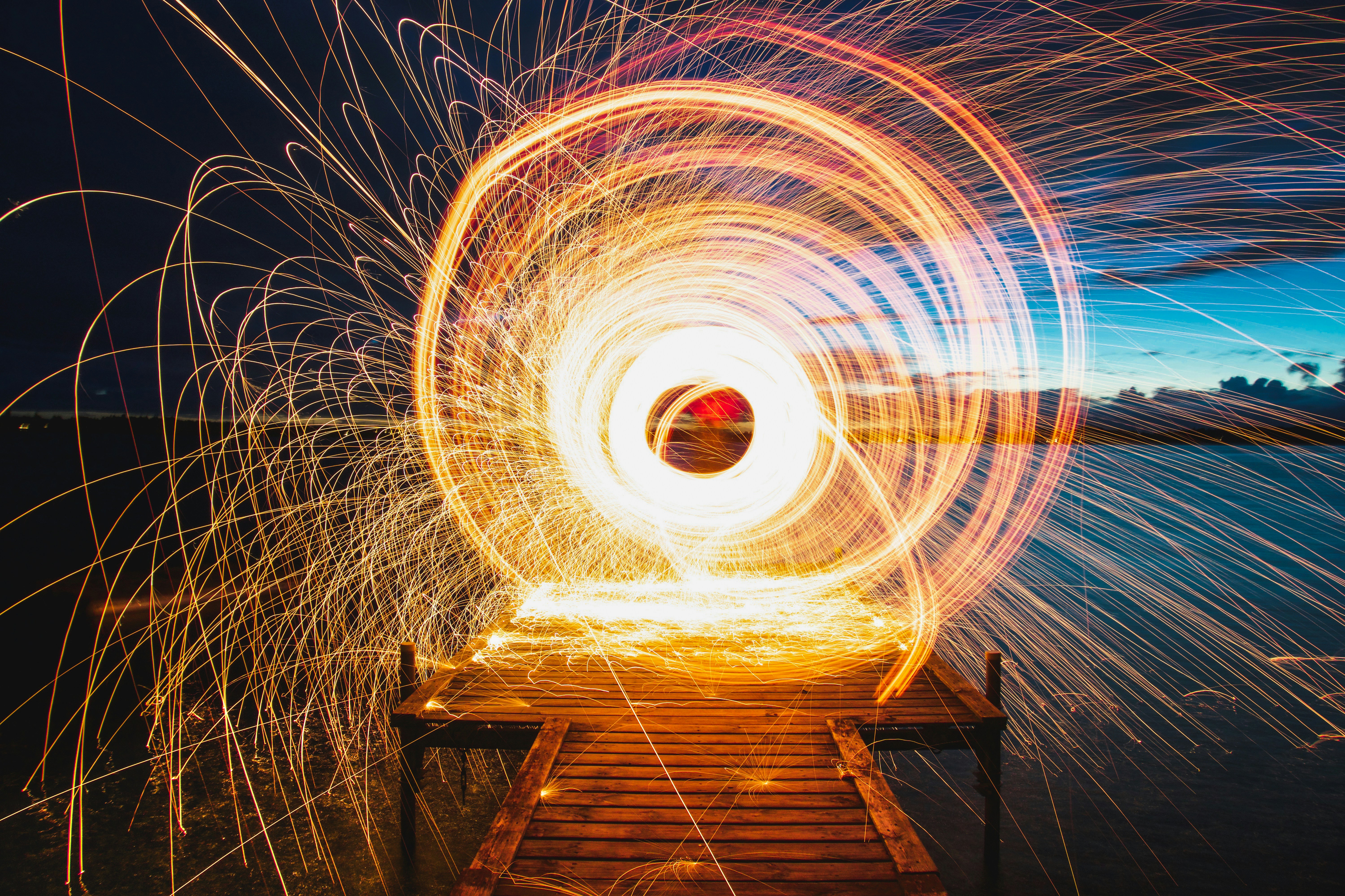Long exposure of swirling steel wool sparks on a wooden pier at dusk.