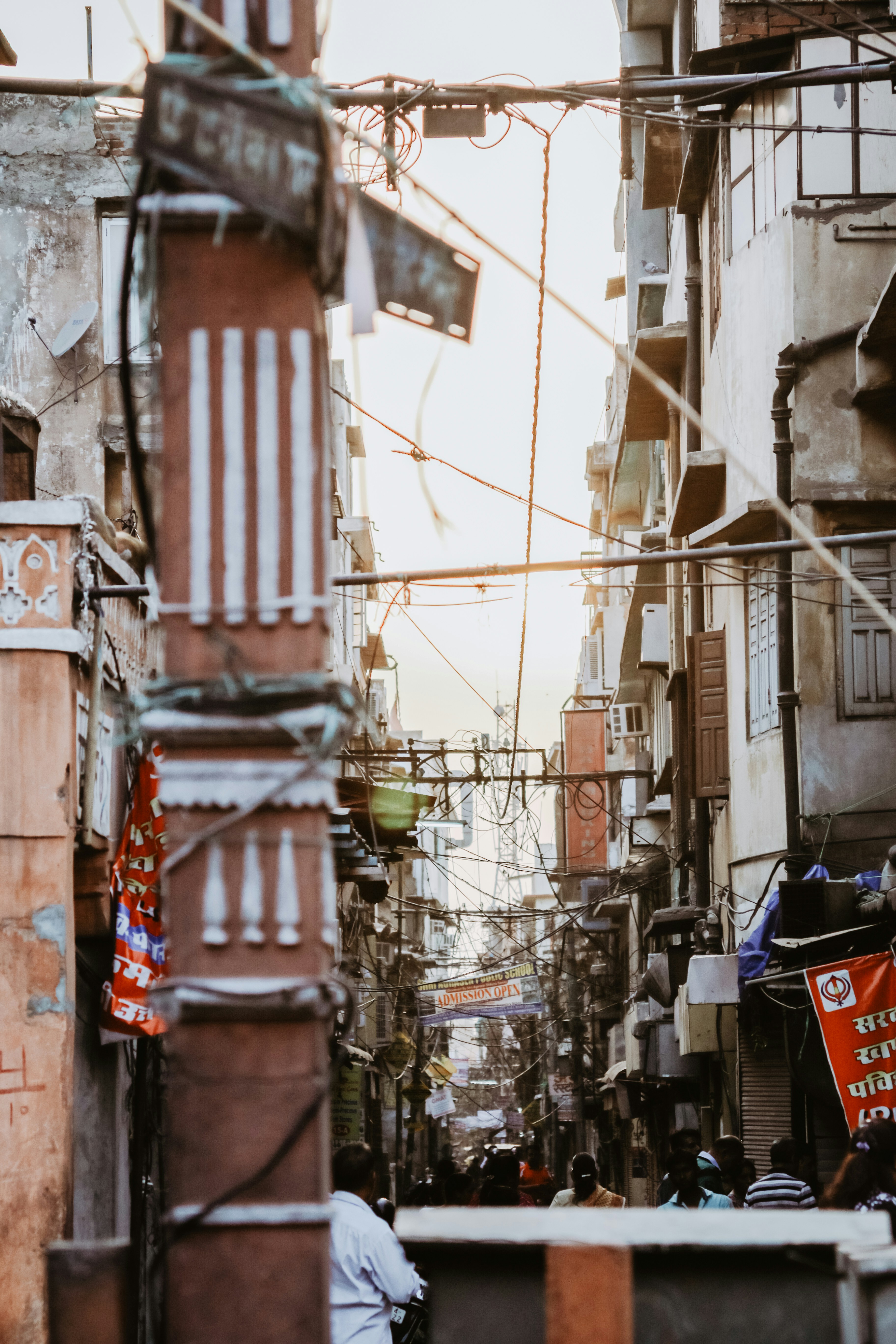 Narrow alleyway bustling with activity, framed by towering buildings and tangled power lines. Colorful banners add vibrancy to the scene.