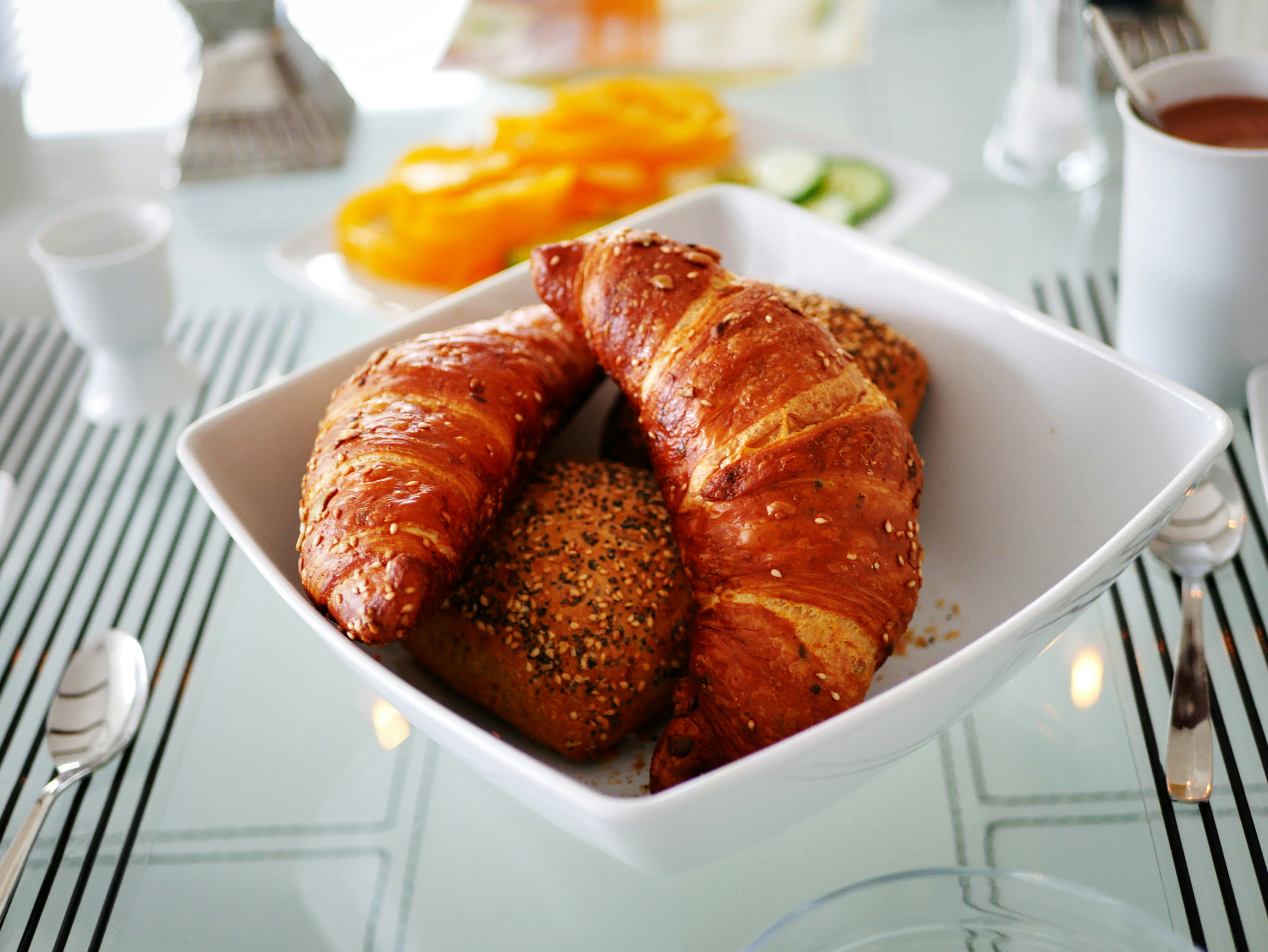 Golden croissants and a seeded roll in a white bowl on a reflective breakfast table.