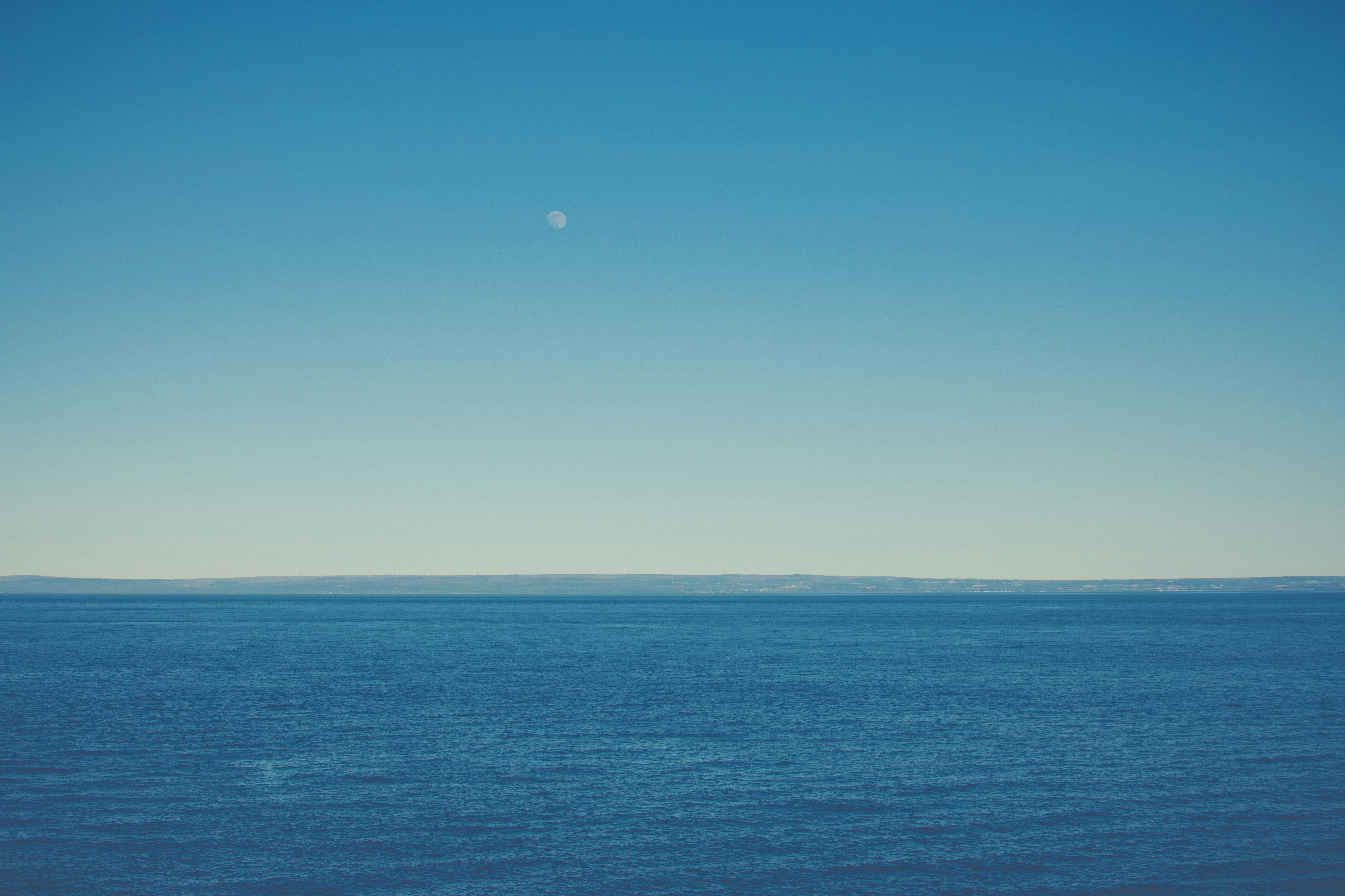 Expansive view of a tranquil sea under a clear blue sky with a solitary moon in the distance.