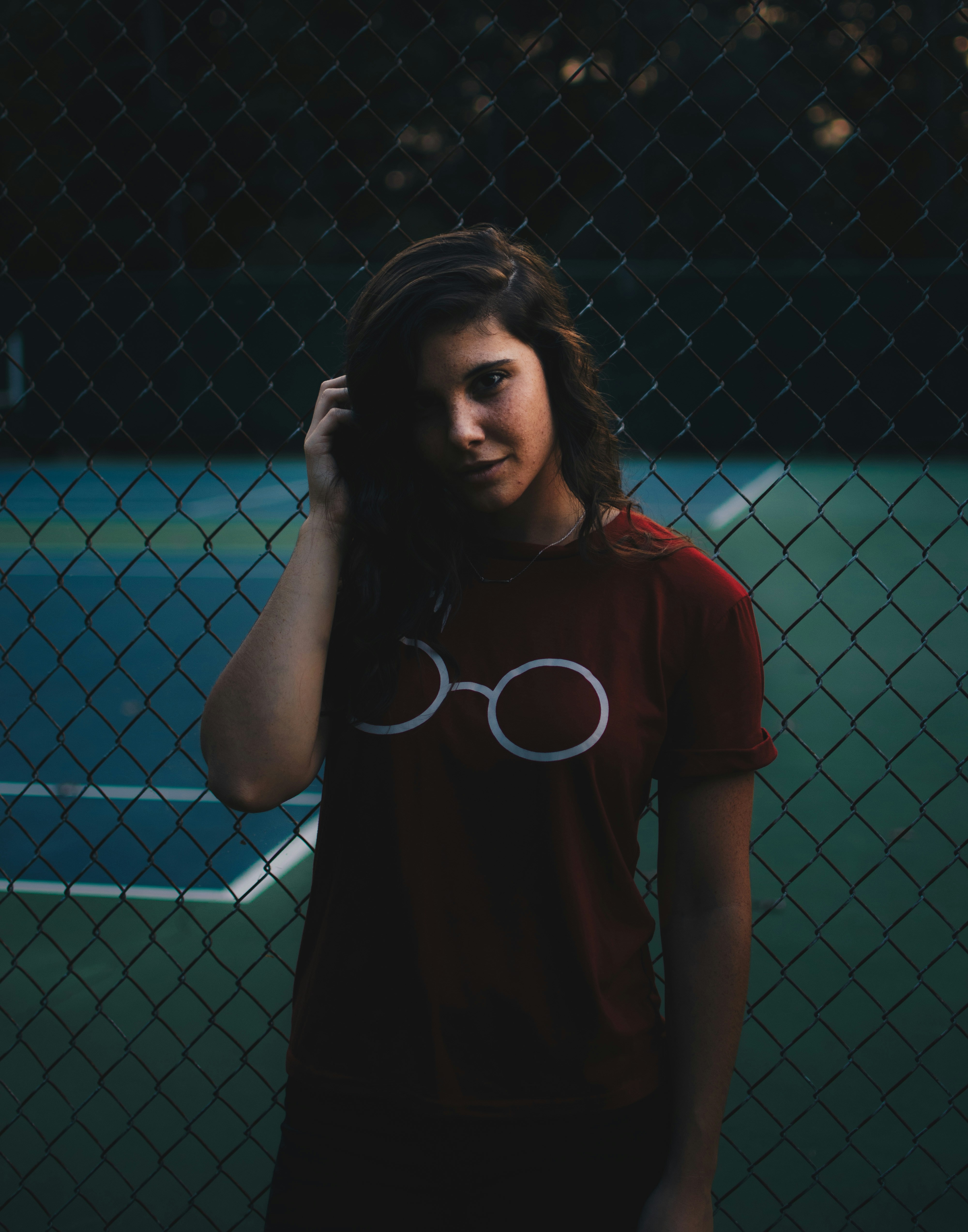 A young woman stands against a chain-link fence on a tennis court, her expression contemplative as she lightly touches her hair.