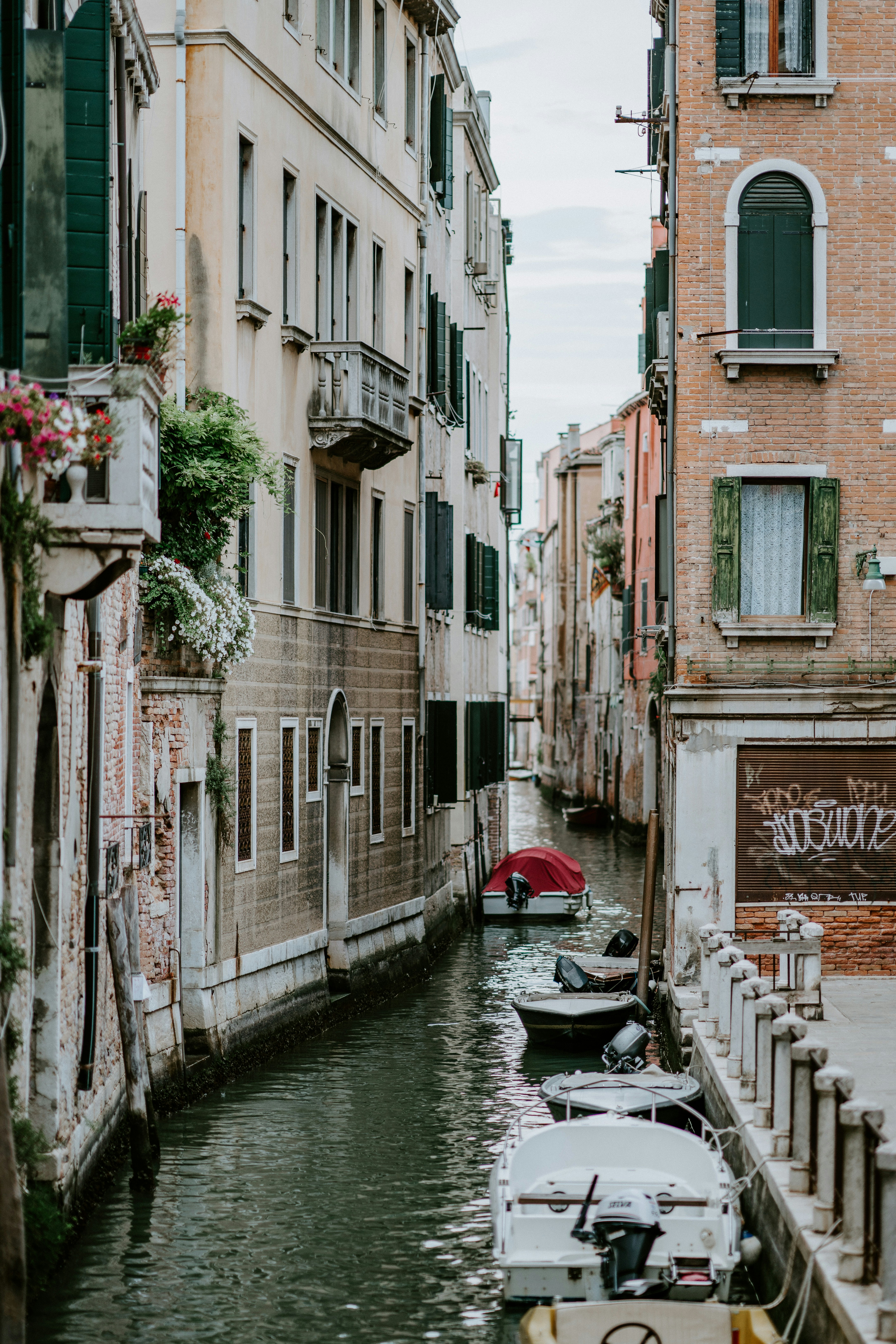 Narrow canal in Venice flanked by charming buildings, with colorful boats moored along the banks. Lush greenery adds a touch of vibrancy.