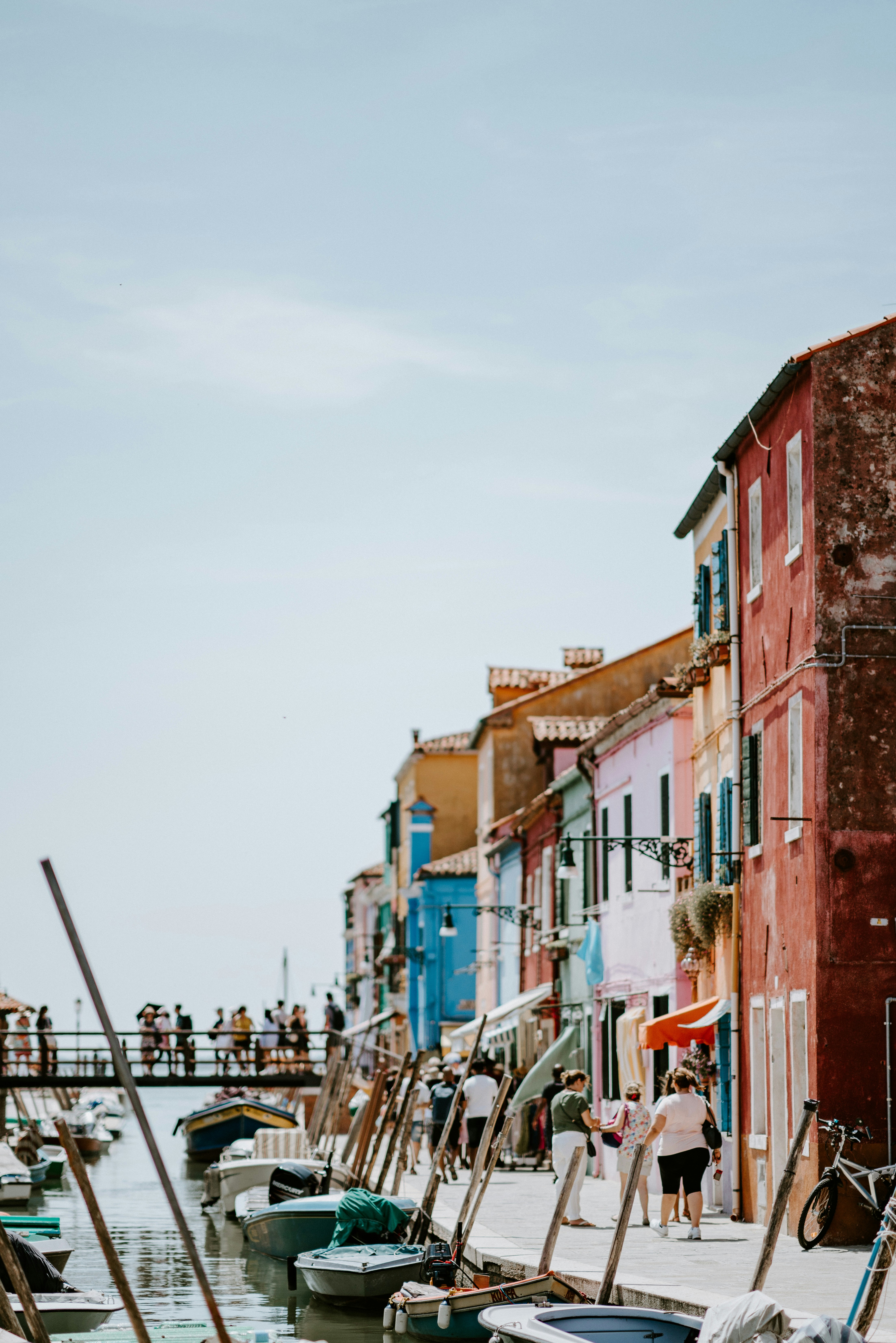 This captivating image showcases a lively canal bordered by a row of vividly painted buildings in hues of red, blue, and yellow, epitomizing the charm of a picturesque European town. The soft, natural lighting enhances the bright colors and creates a serene atmosphere, while pedestrians and boats add a dynamic element to the scene. The composition captures the essence of everyday life against a backdrop of architectural beauty, making it a visually striking portrayal of culture and color.