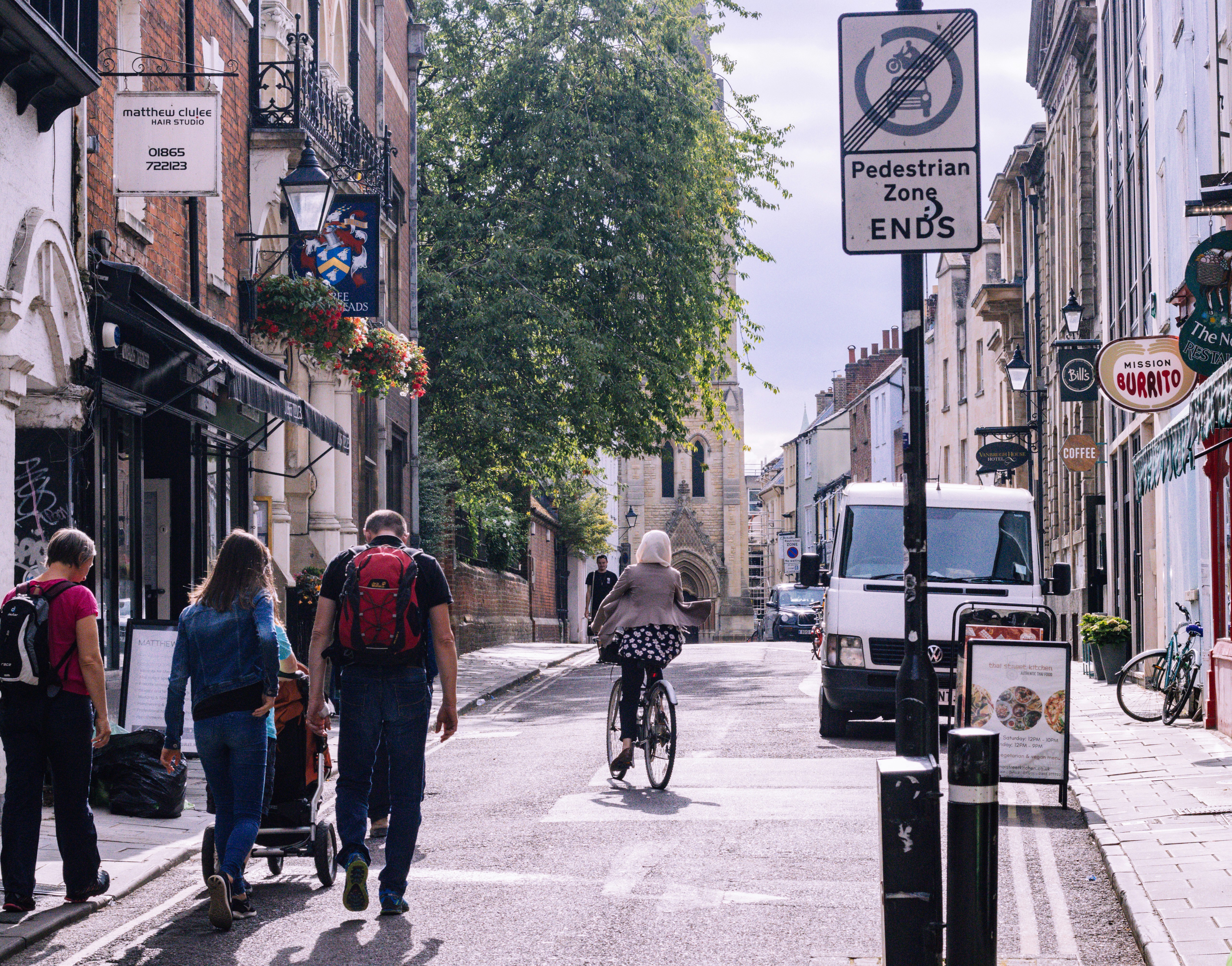 People walking and cycling down a quaint, sunlit street flanked by historic buildings.