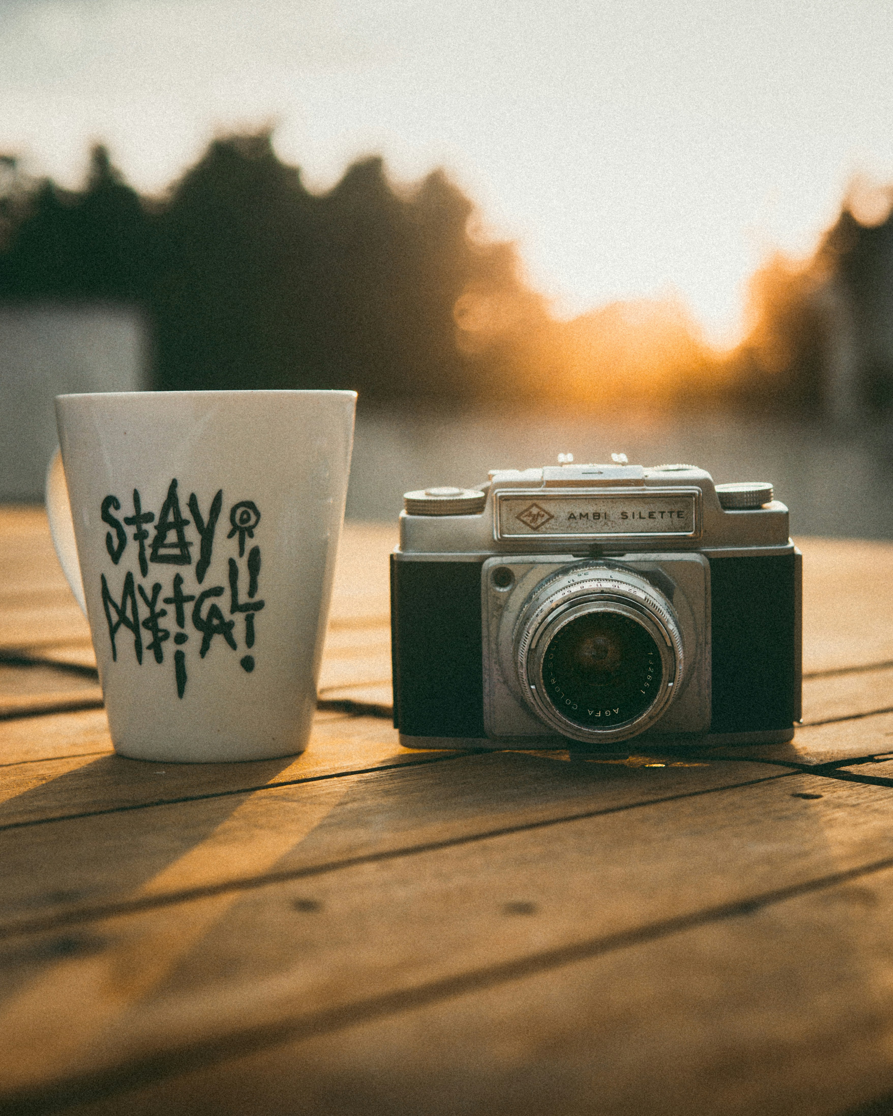 Vintage camera and a coffee mug with a playful design on a wooden table at sunset.