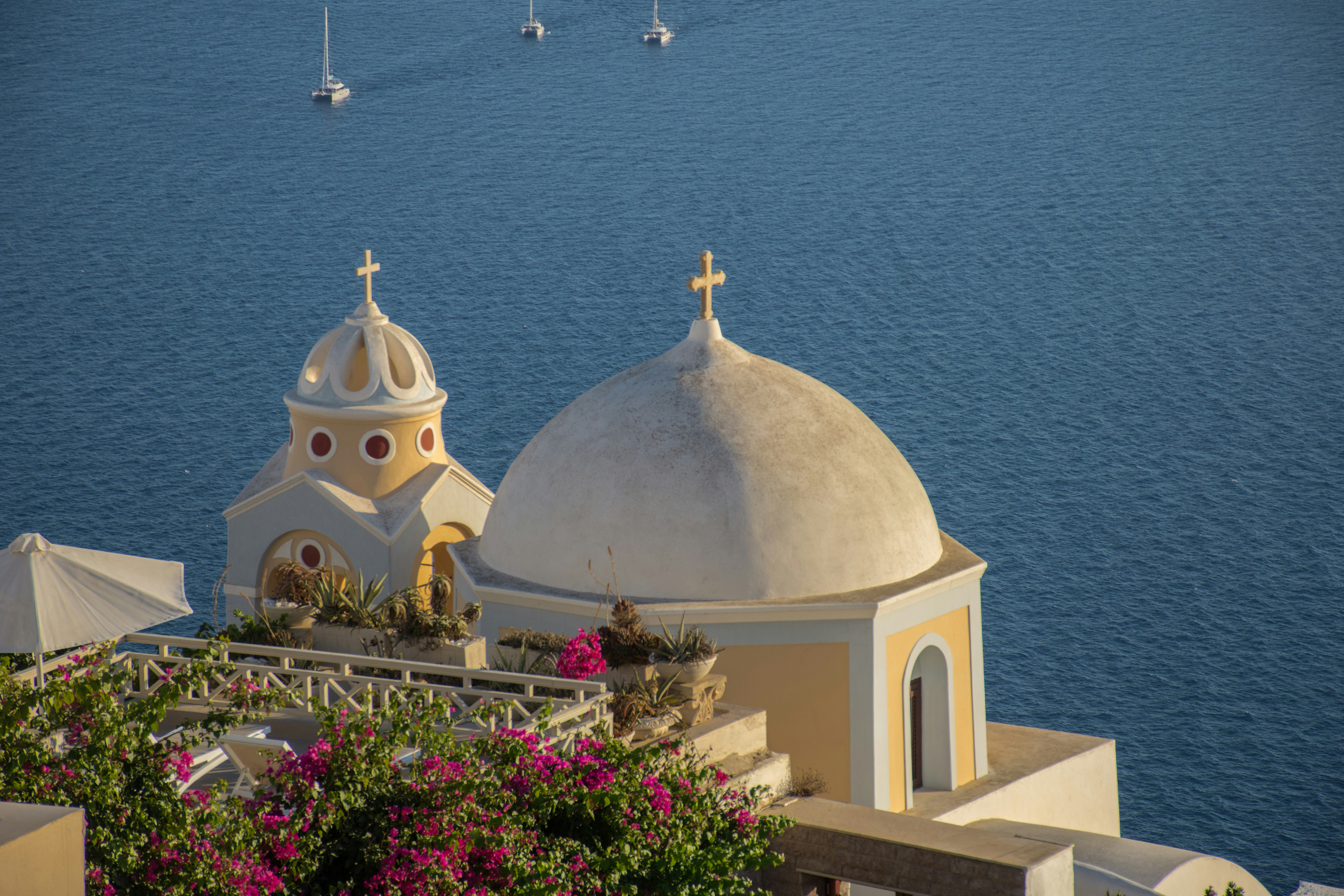 Sunlit domed church overlooking the blue Aegean Sea with scattered sailboats.