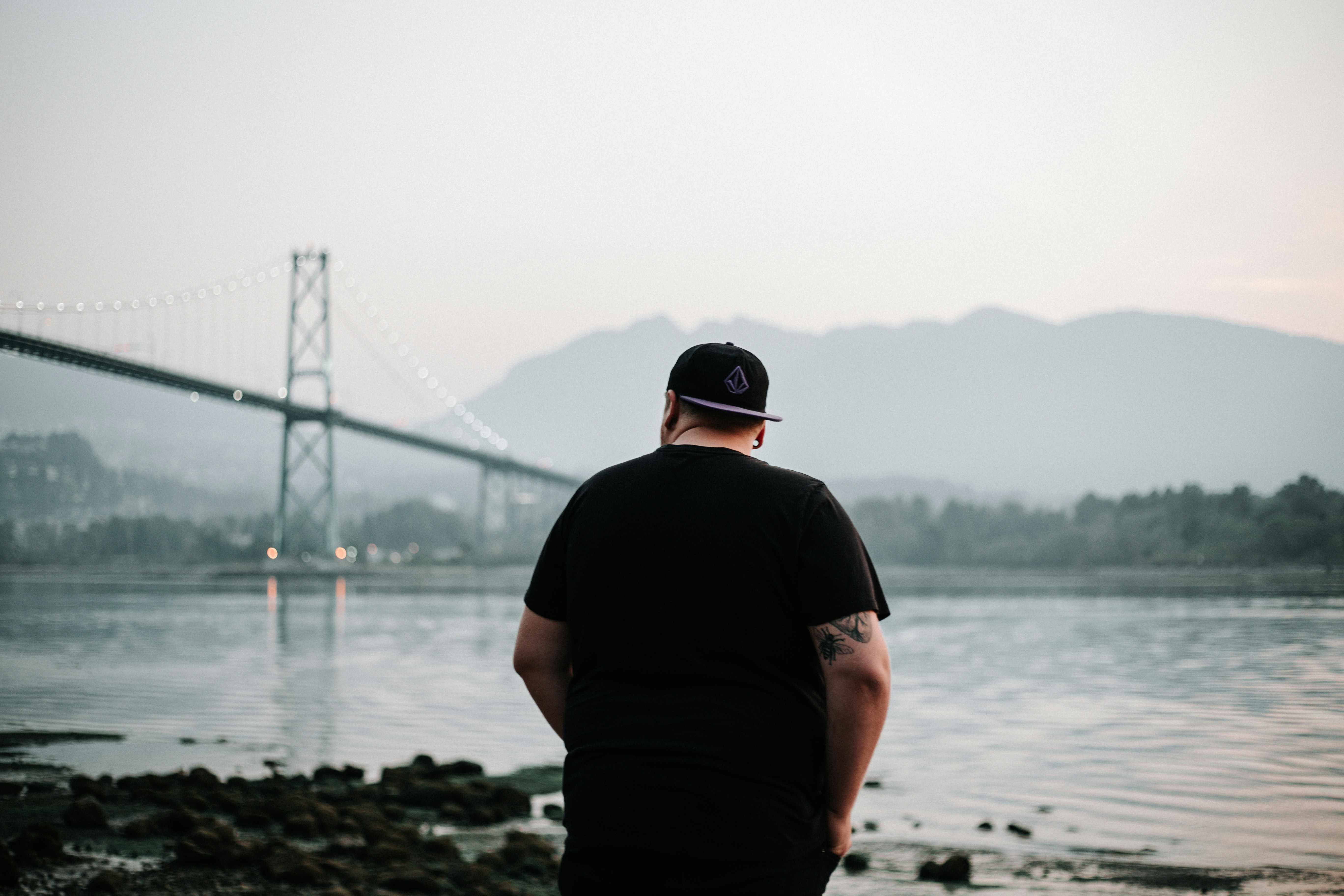 Silhouetted figure gazing over calm waters with a bridge in the background at dusk. The scene conveys a sense of tranquility and introspection.