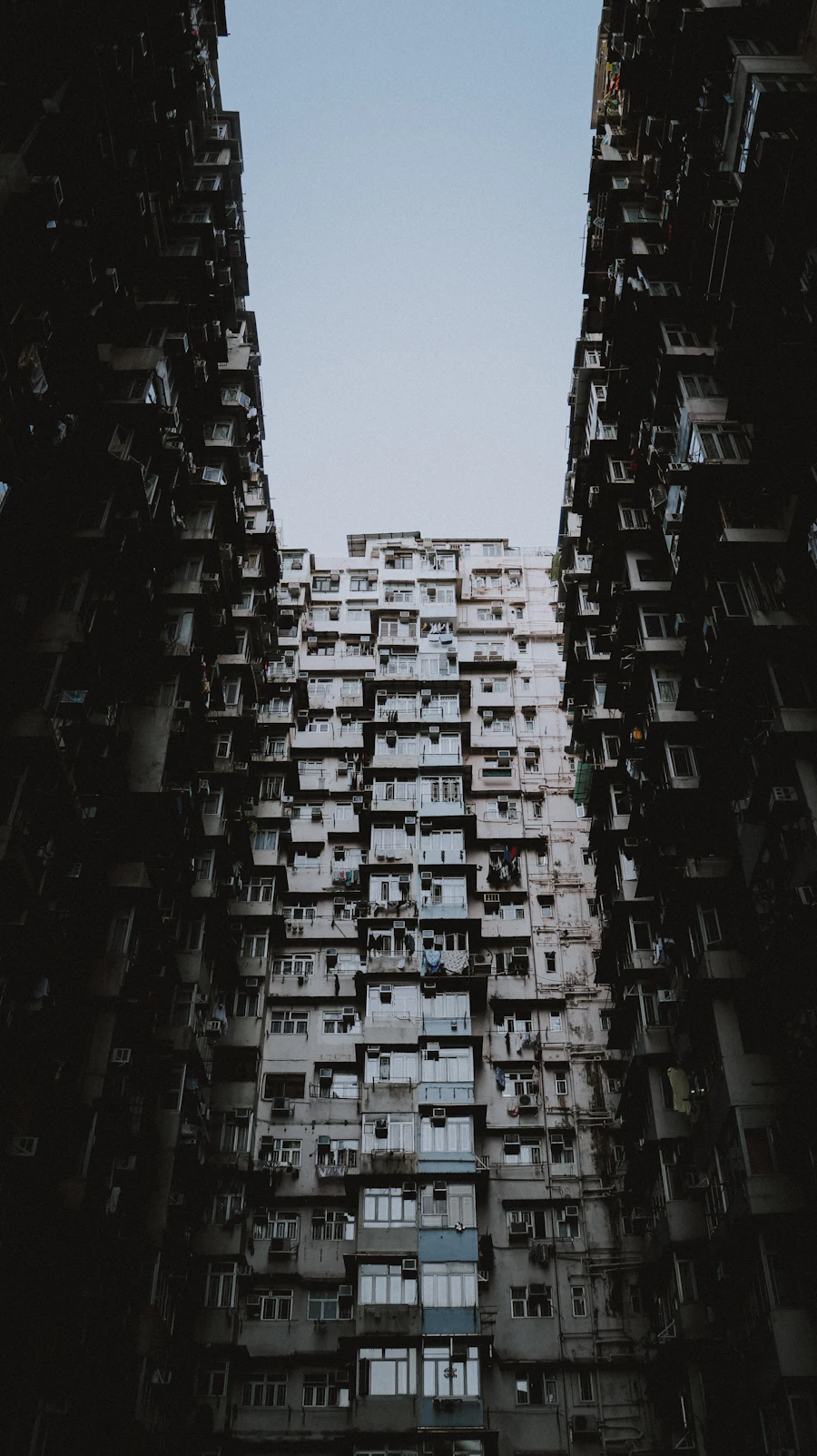 A high-rise courtyard view with two dark apartment blocks framing a weathered residential tower and a sliver of pale sky above. Dense rows of windows and balconies create a stark urban canyon.
