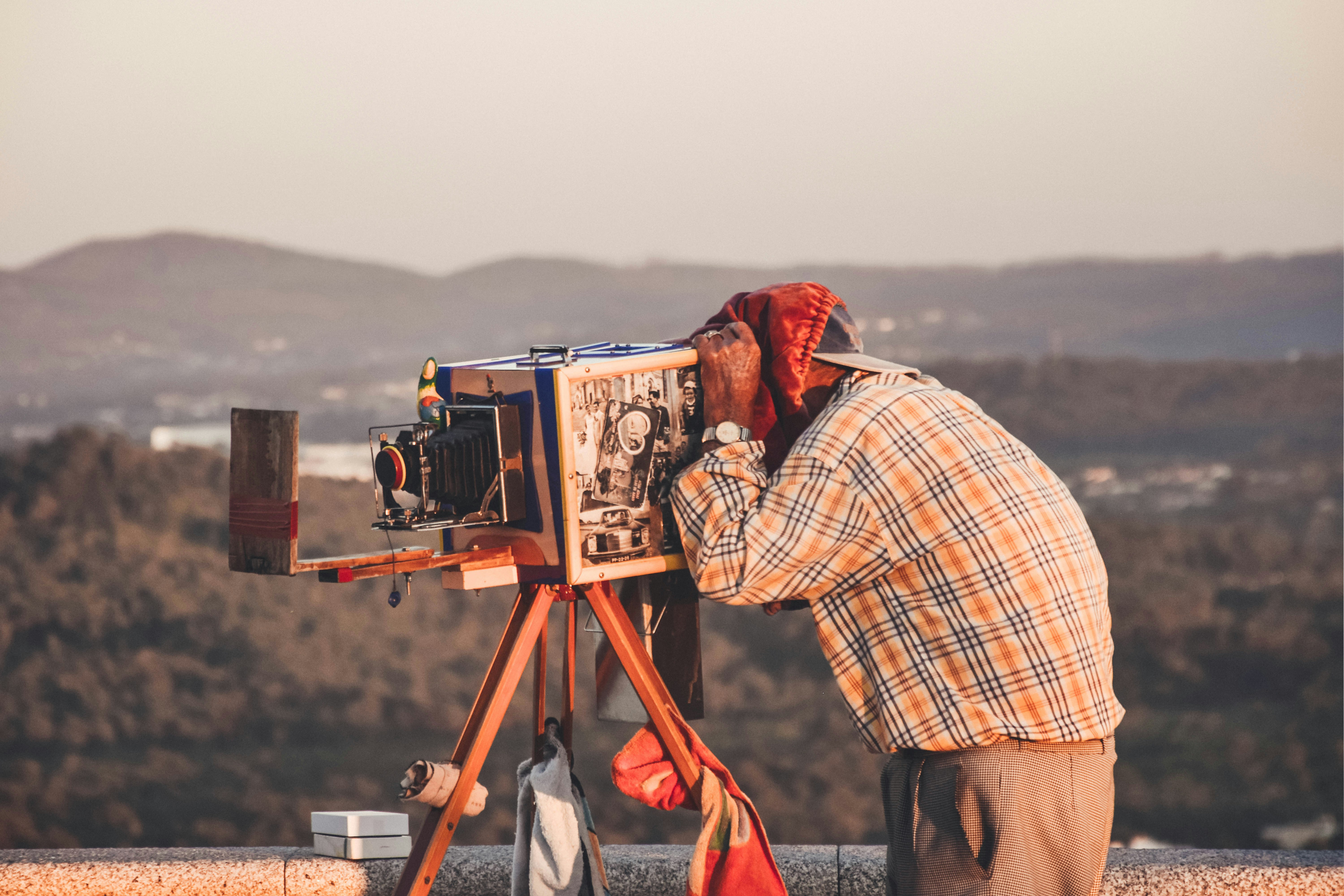 Person using a vintage folding camera on a tripod against a mountainous backdrop at sunset.