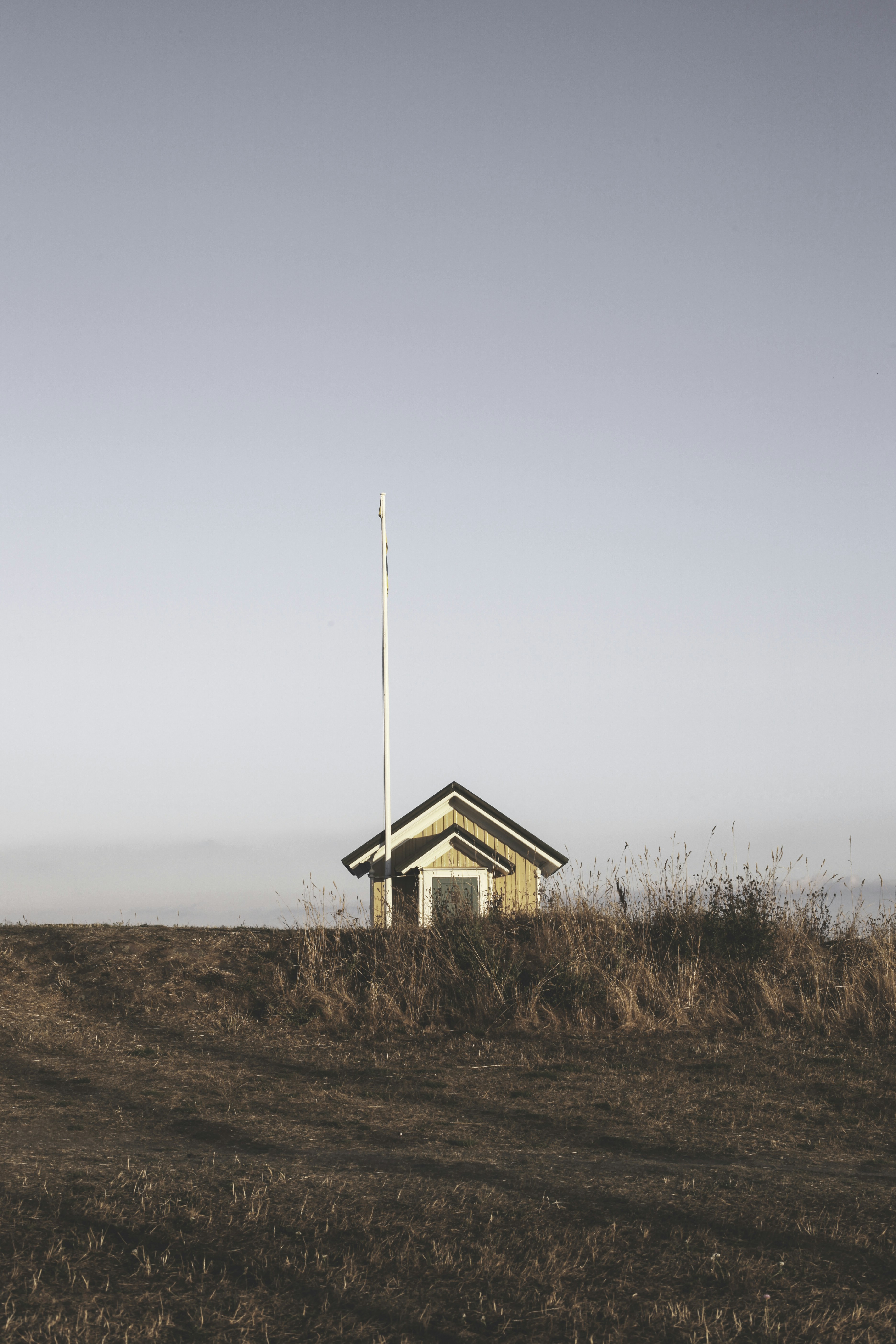 A quaint yellow house stands alone on a grassy hill, with a flagpole rising beside it against a soft, muted sky.