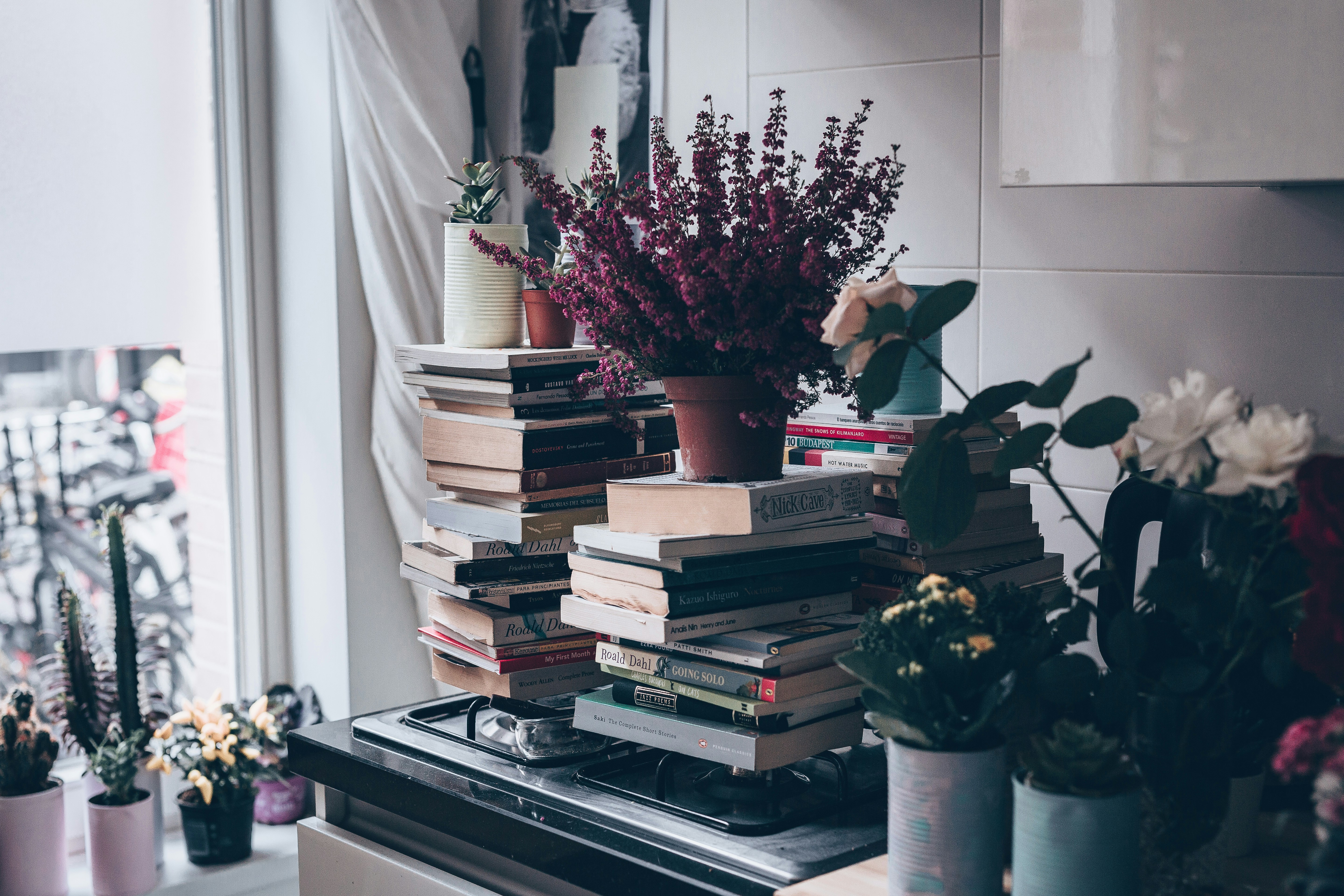 A vibrant arrangement of stacked books crowned by a blooming pot of purple flowers, surrounded by potted plants in a cozy kitchen setting.