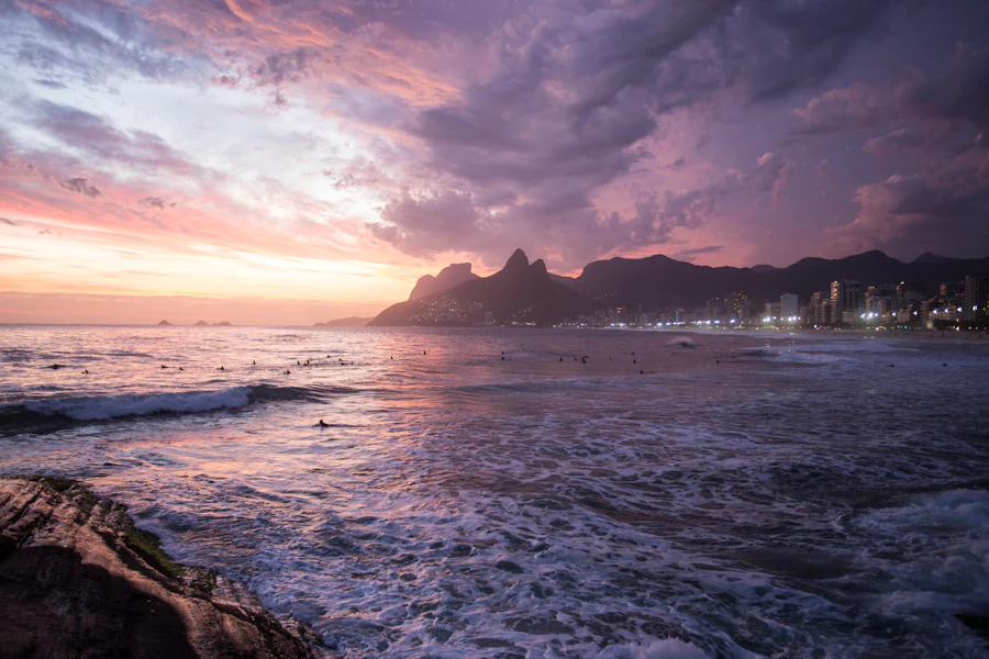 Surfers catching waves under vibrant pink and purple sunset skies near mountains and city lights.