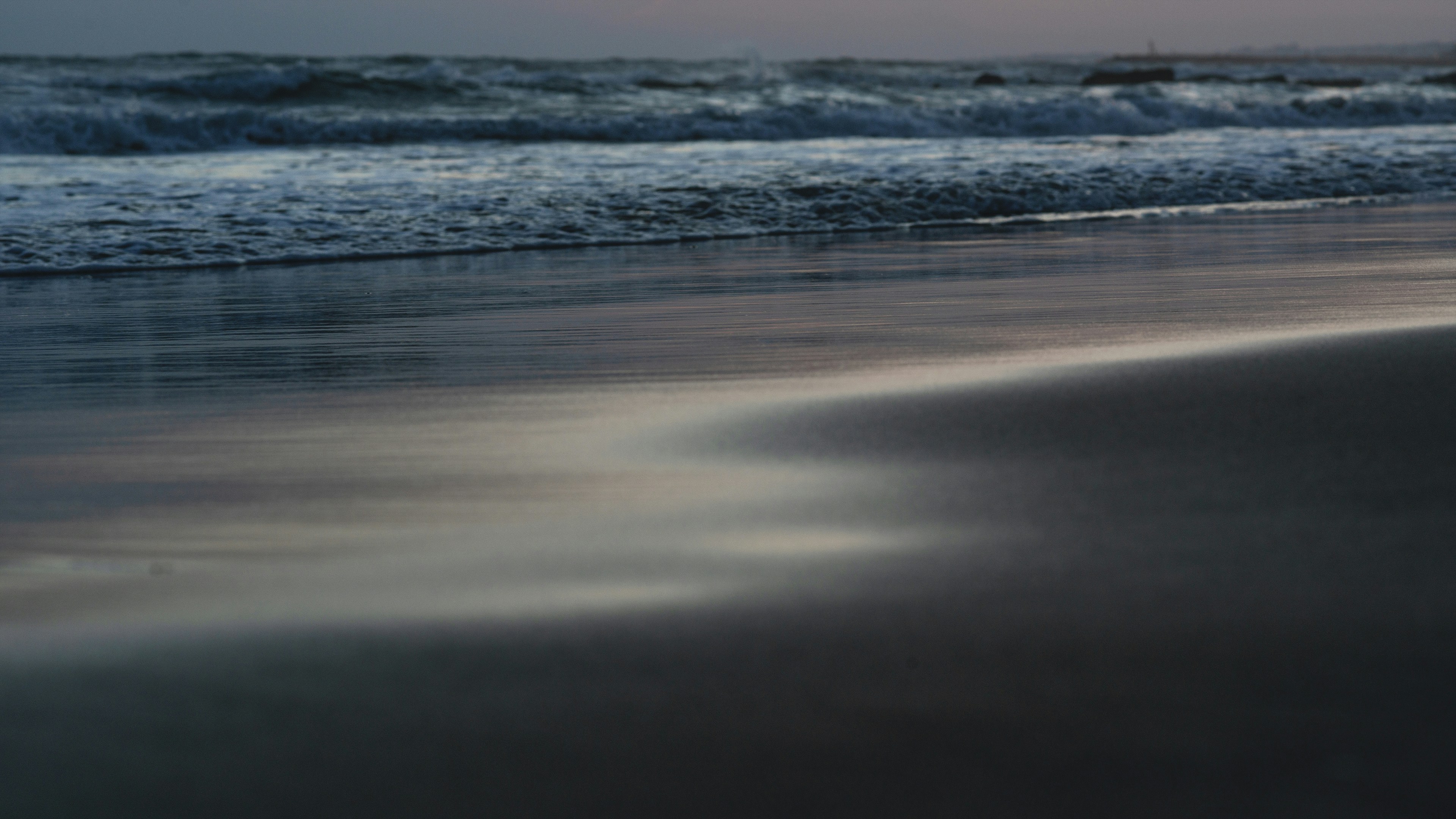 Gentle ocean waves caress a dark sandy shore under a dimly lit evening sky.