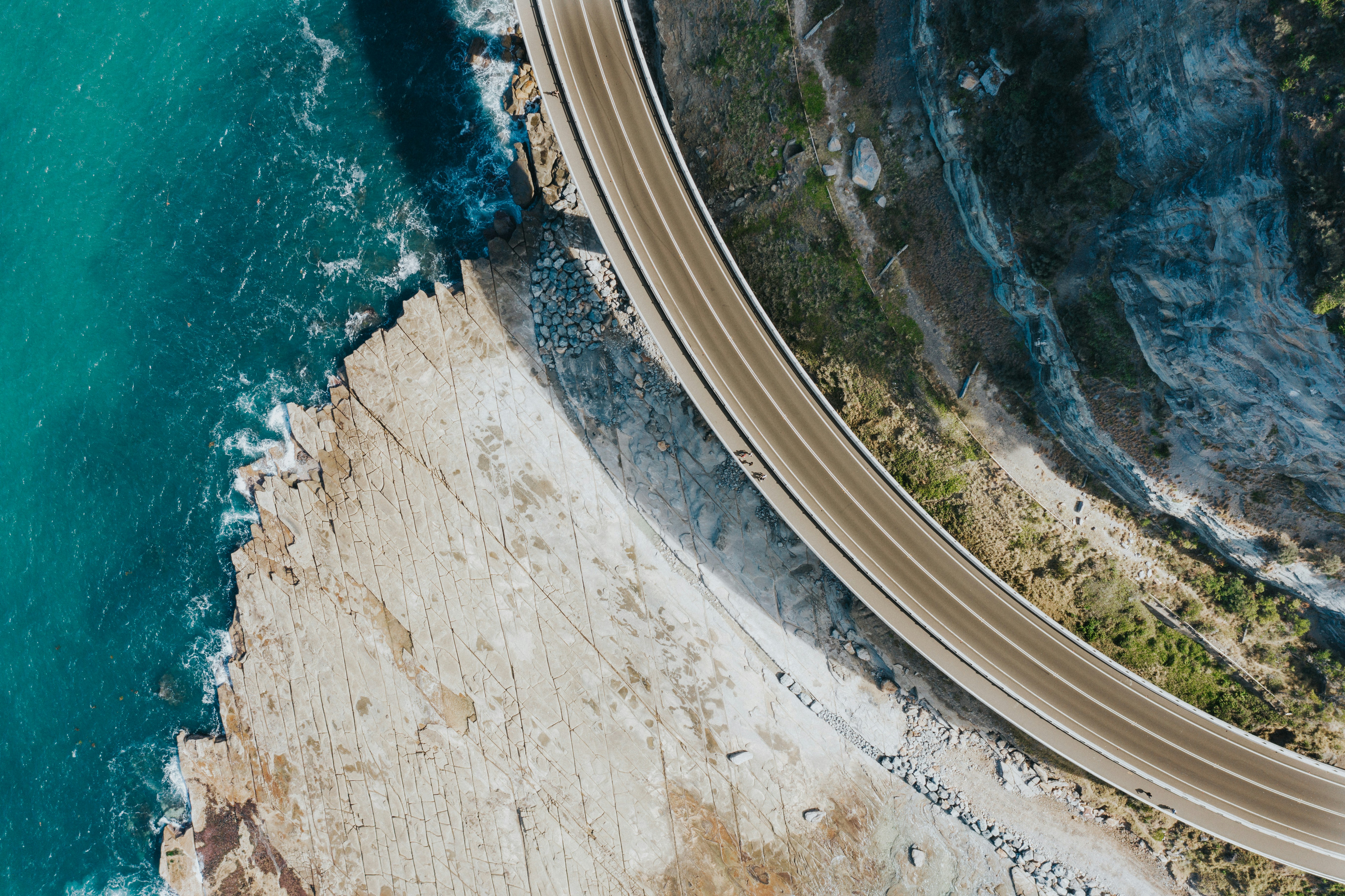 Aerial view of a winding road curving along a rocky coastline, juxtaposed against the vibrant blue ocean. The landscape showcases the interplay between natural and human-made elements.