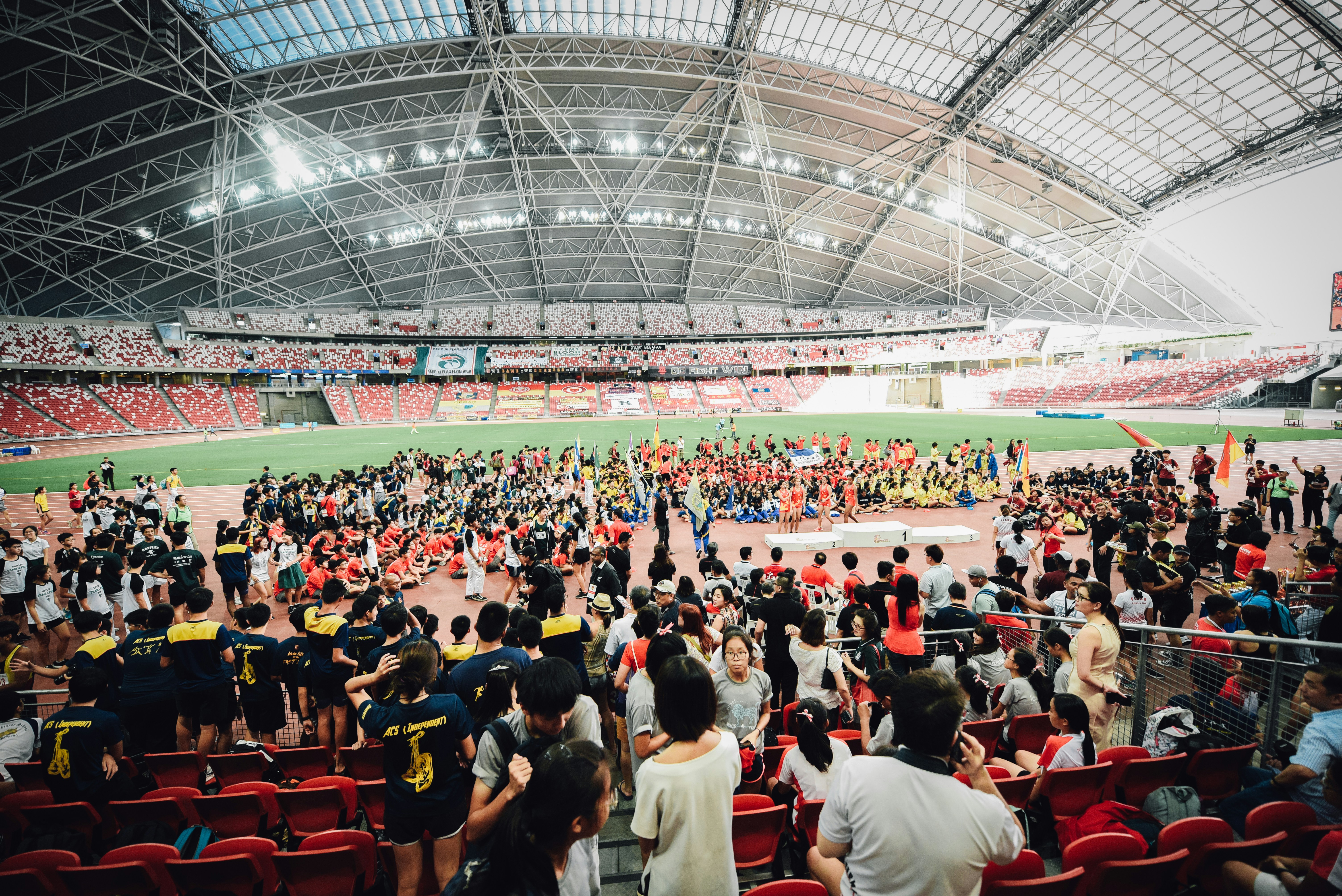 A lively crowd fills a stadium during an event, showcasing a variety of colorful outfits and enthusiastic participants. The architectural design of the dome looms overhead.