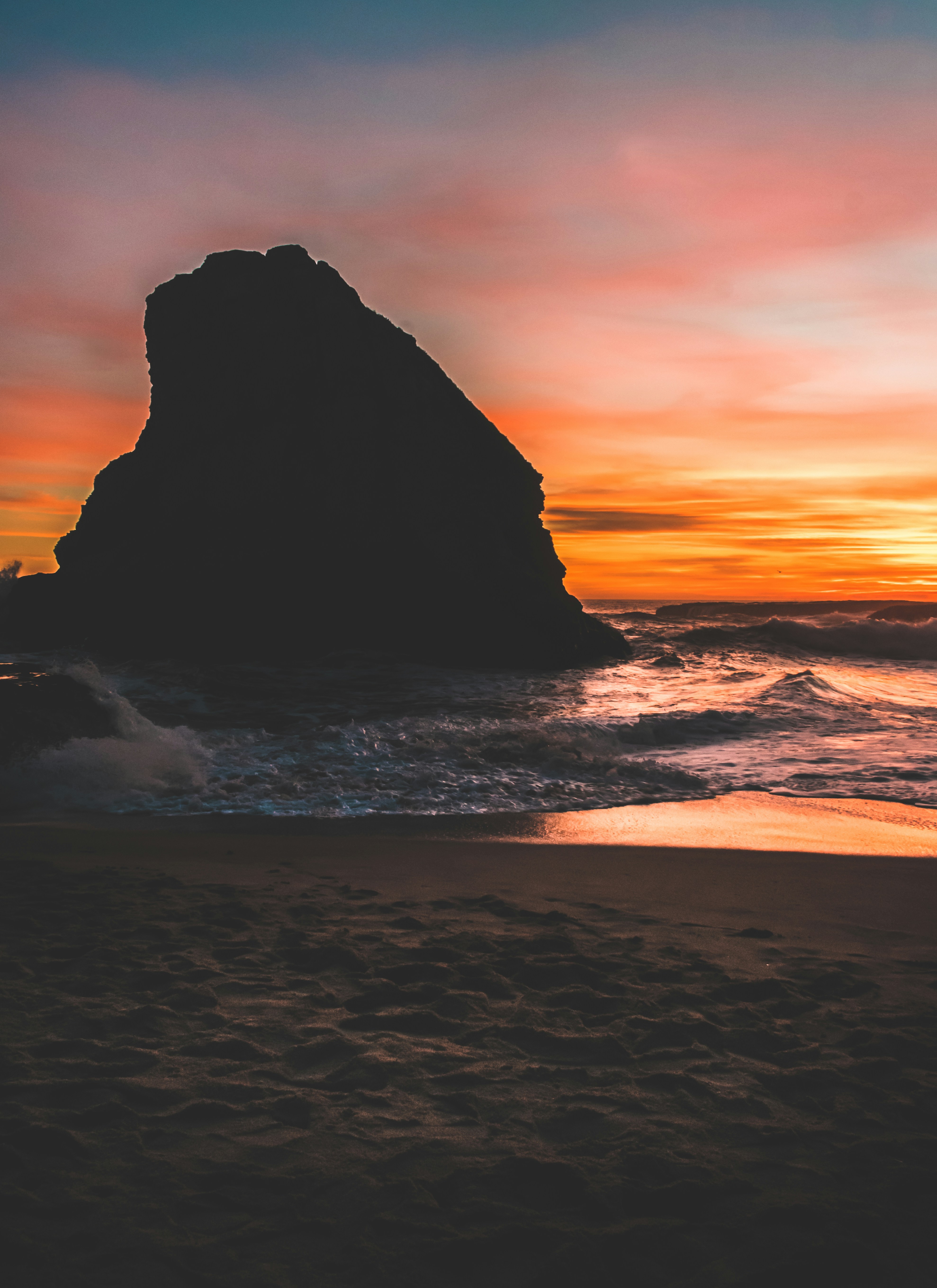 A large rock formation silhouetted against a colorful sunset, with gentle waves lapping at the shore. The scene captures the tranquil beauty of nature.