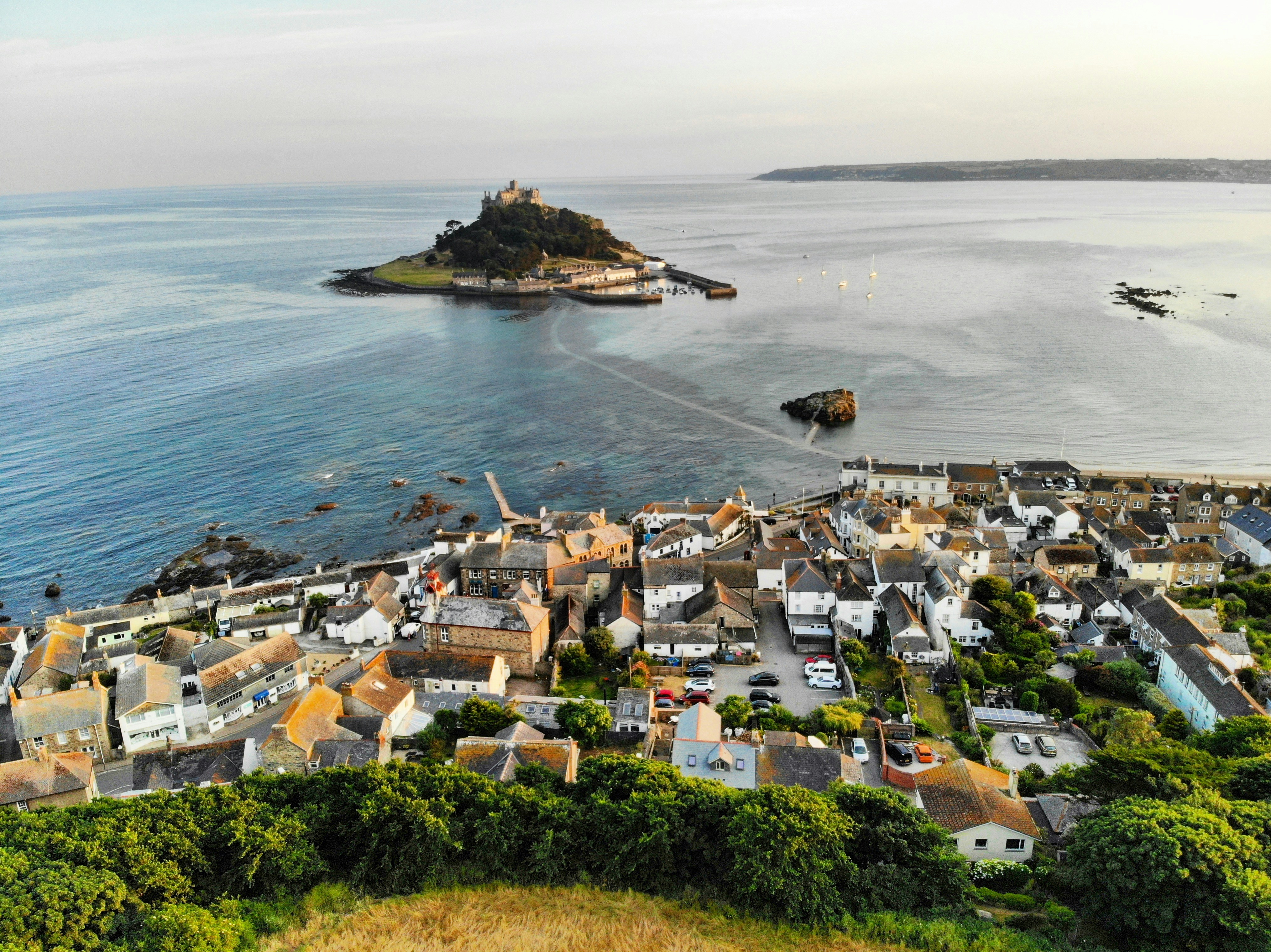 Aerial view of a coastal village with quaint houses leading to a historic castle on an island, surrounded by calm waters.
