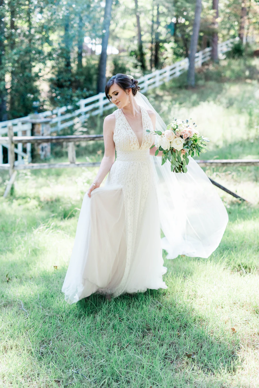Bride in a lace gown holding a bouquet and veil in a bright wooded meadow with a white fence behind her.