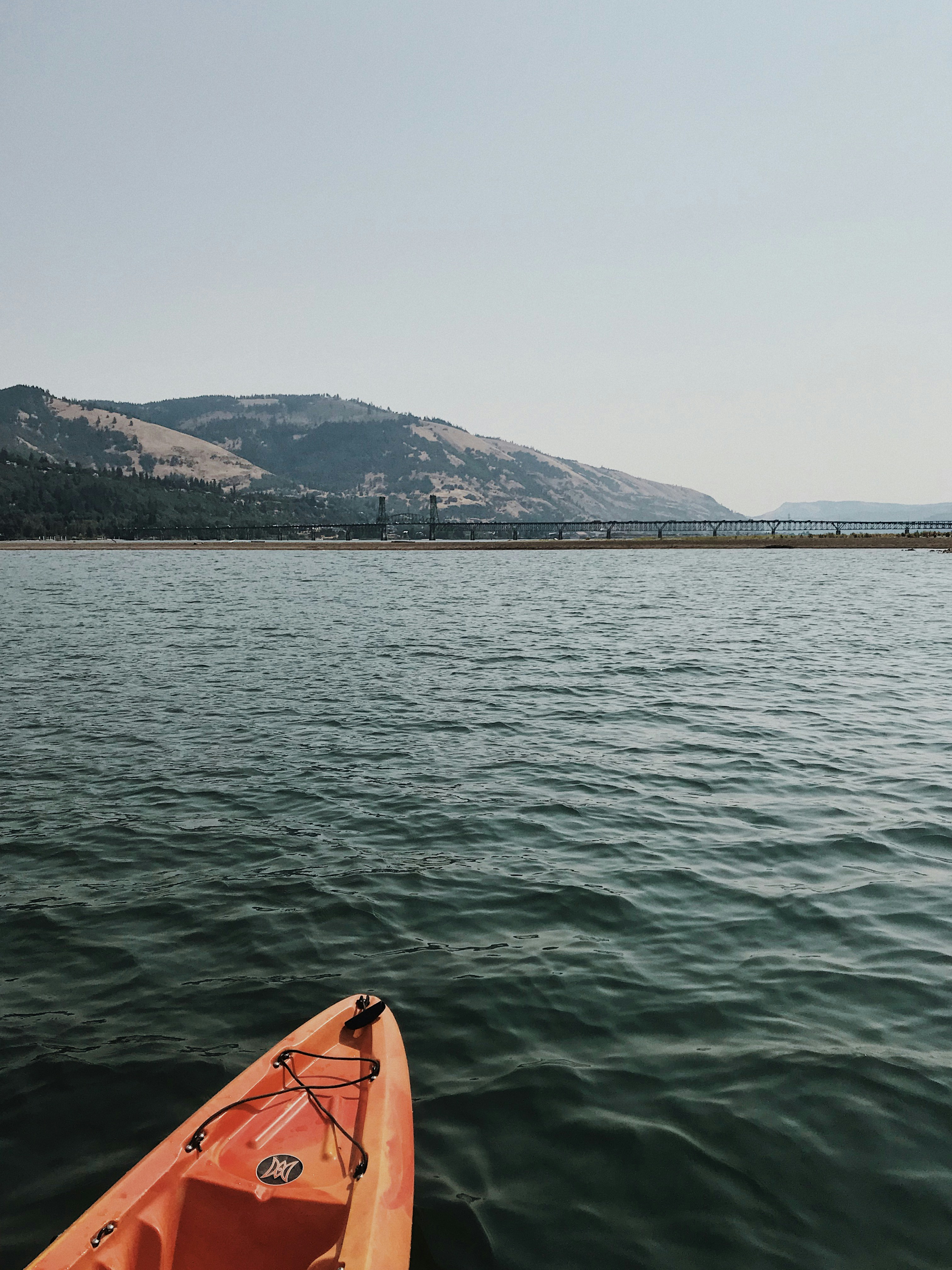 A vibrant orange kayak floats on calm waters, with distant hills and a bridge under a clear sky. The scene evokes a sense of peace and connection with nature.