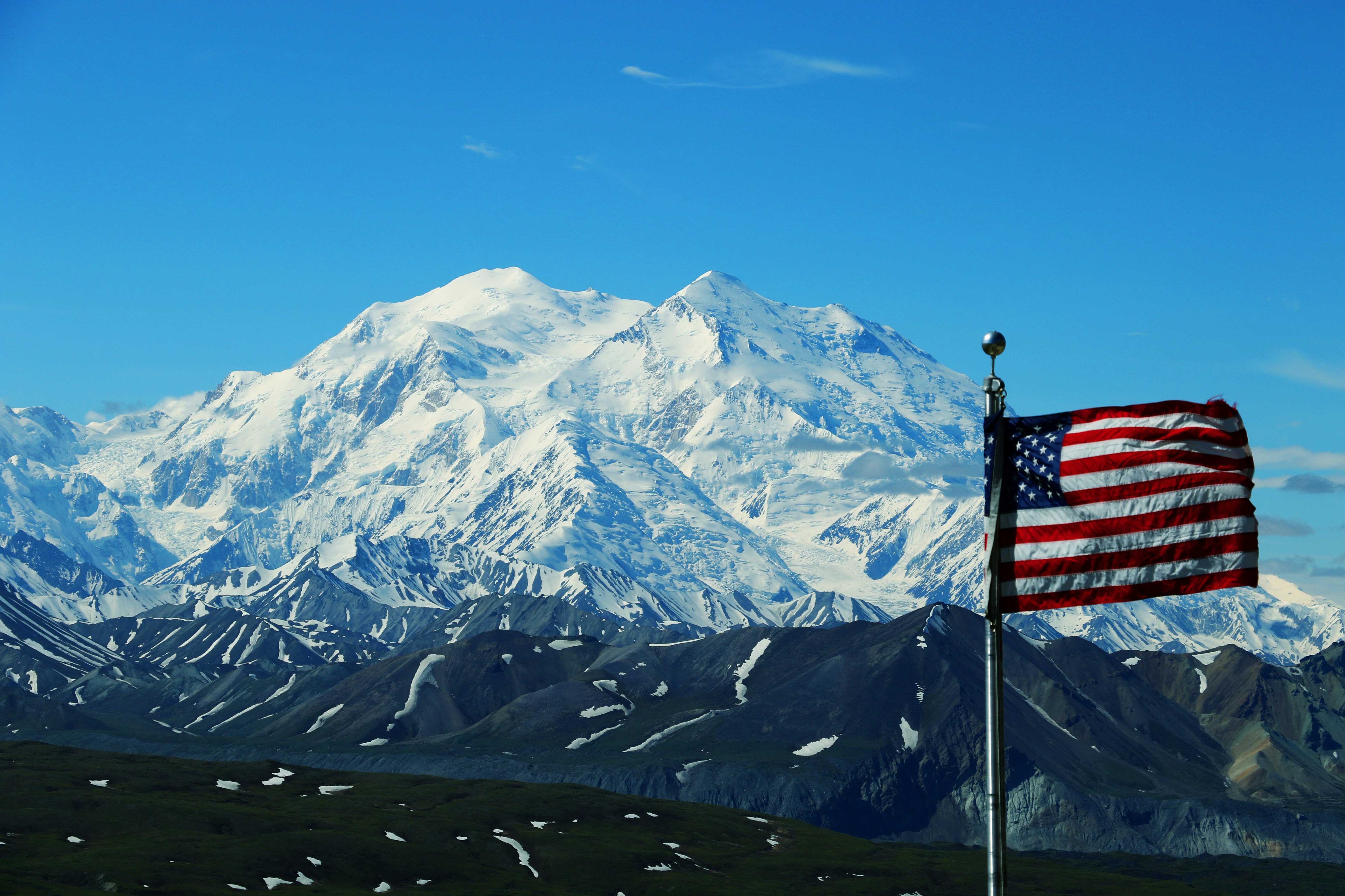 American flag waving in the foreground with a backdrop of snow-capped mountains under a clear blue sky.