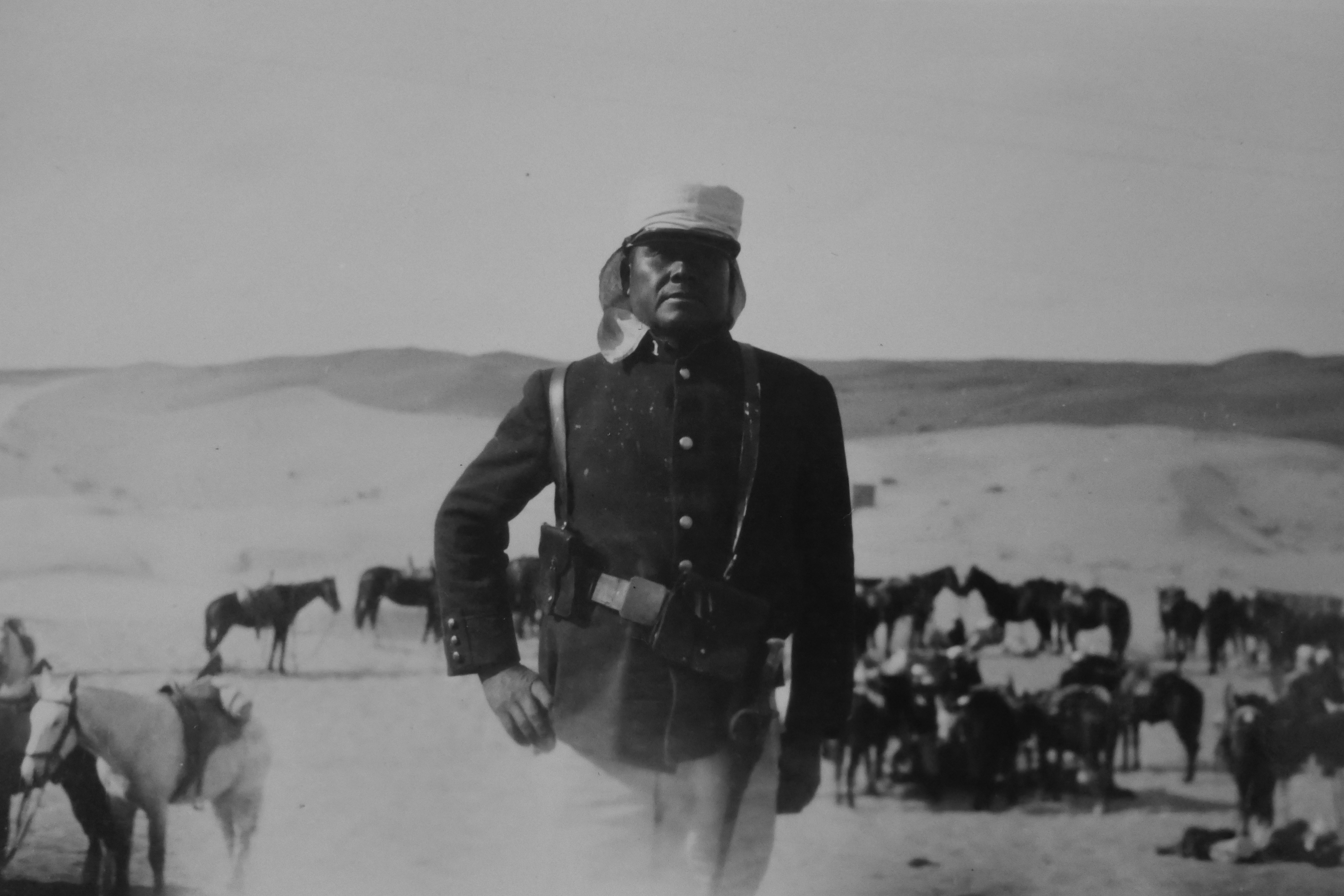 A soldier in historic uniform stands confidently in a desert landscape, surrounded by grazing horses in the background. The scene captures a moment of calm amidst the arid terrain.