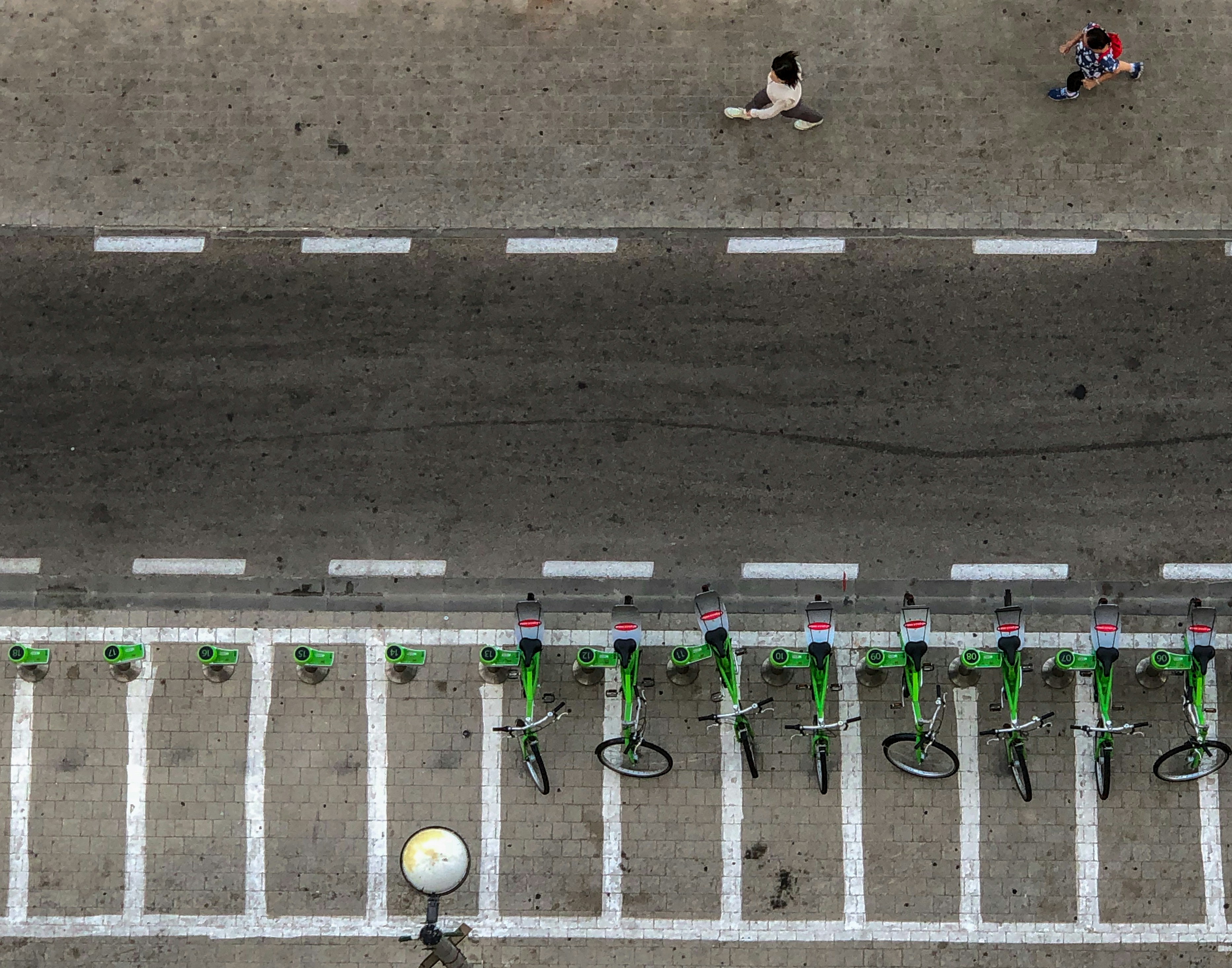 Rows of green bicycles line a city street, viewed from above with pedestrians walking nearby.