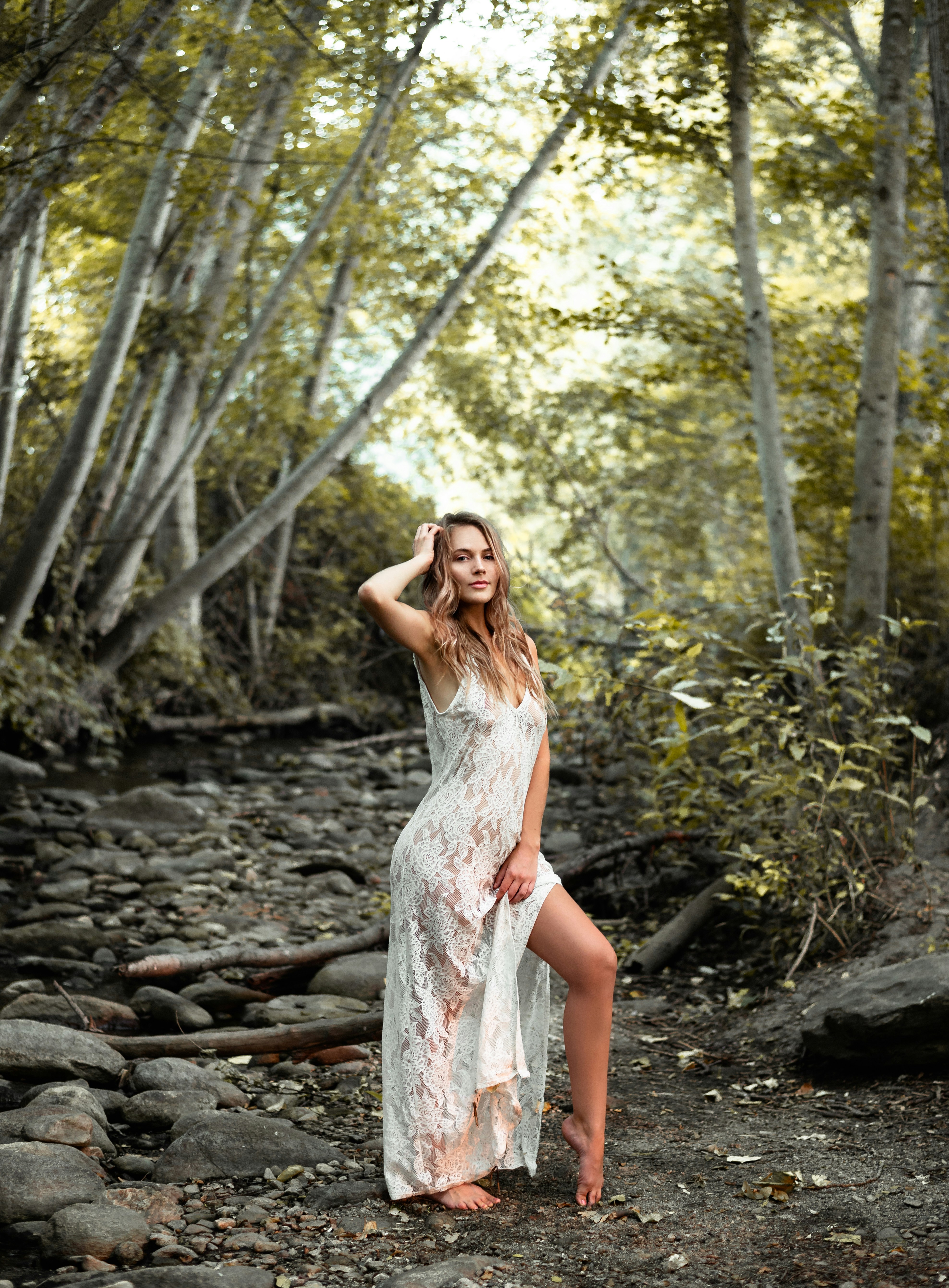 Model in a flowing lace dress poses gracefully among a rocky stream, framed by tall trees and soft, muted colors.