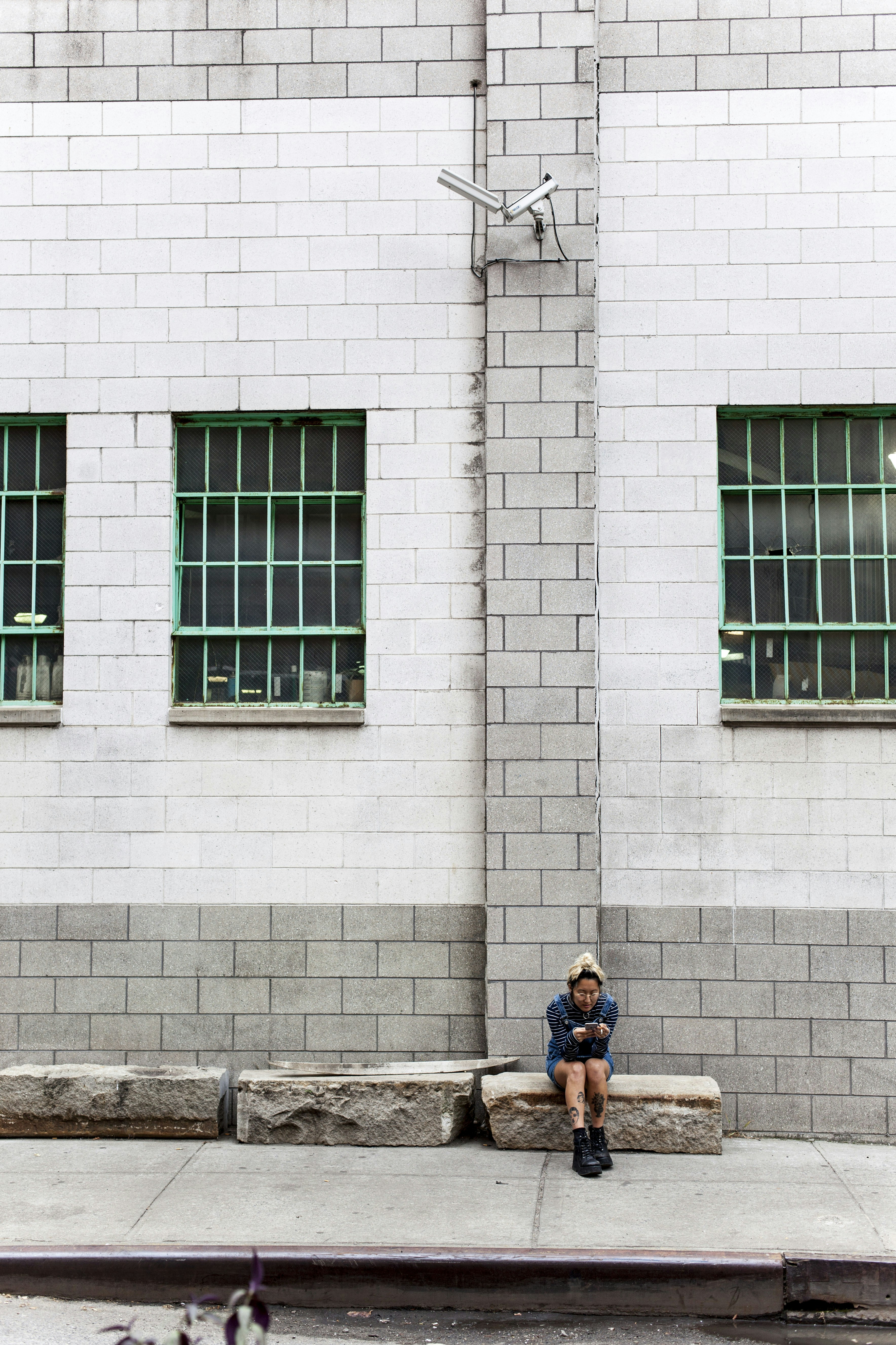 Individual seated on a stone bench against a stark industrial wall, engrossed in a mobile device. Security camera overhead adds a layer of urban narrative.