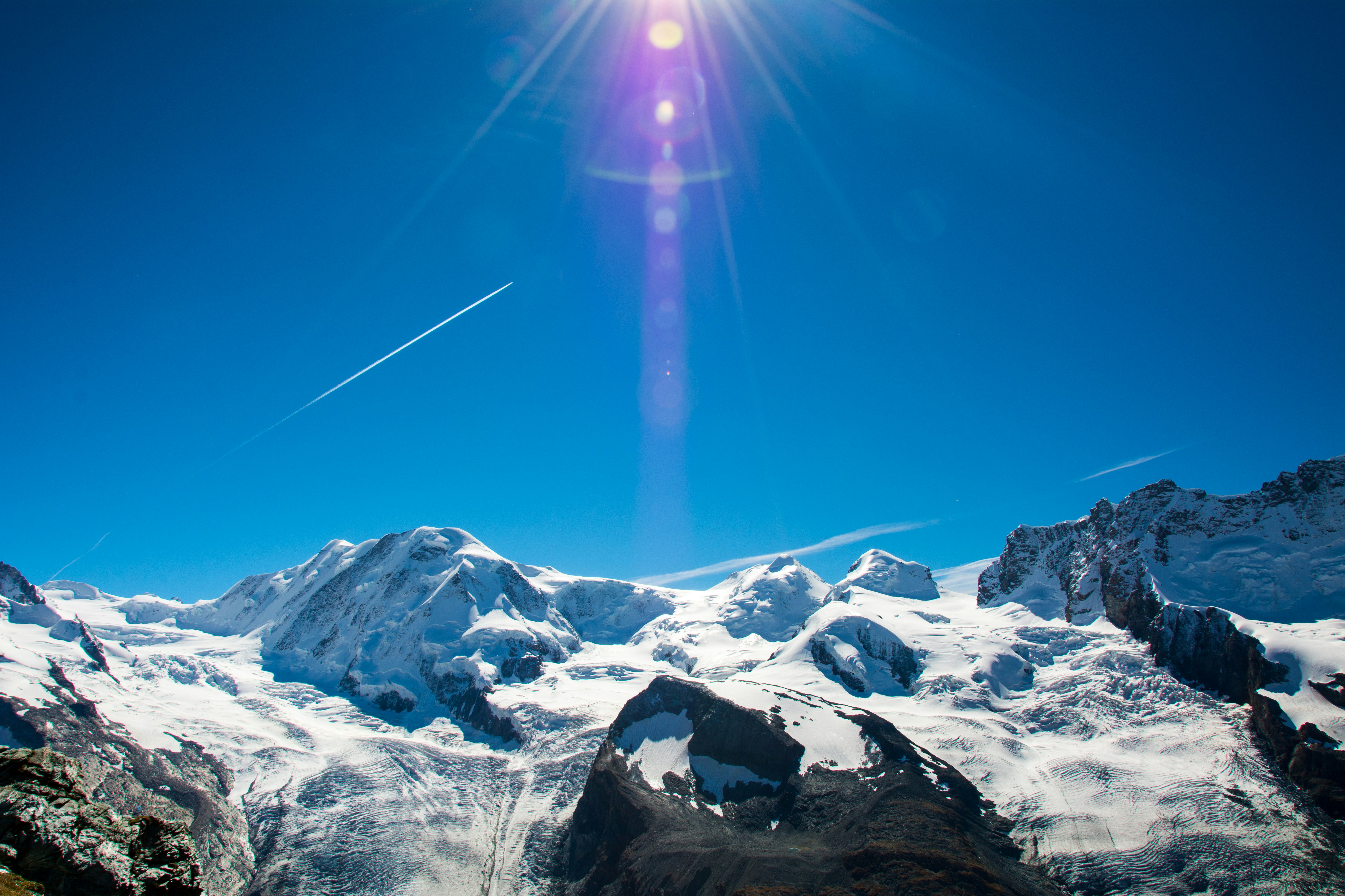 Majestic snow-capped mountains stretch across the horizon, illuminated by a radiant sun against a clear blue sky. Contrails crisscross the atmosphere above.