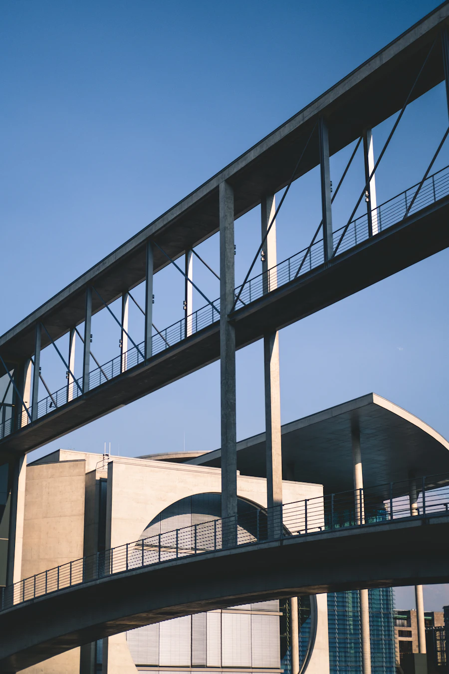 Layered concrete pedestrian bridges and a modern building facade intersect beneath a clear blue sky. Strong diagonal lines and repeating supports create a clean urban composition.