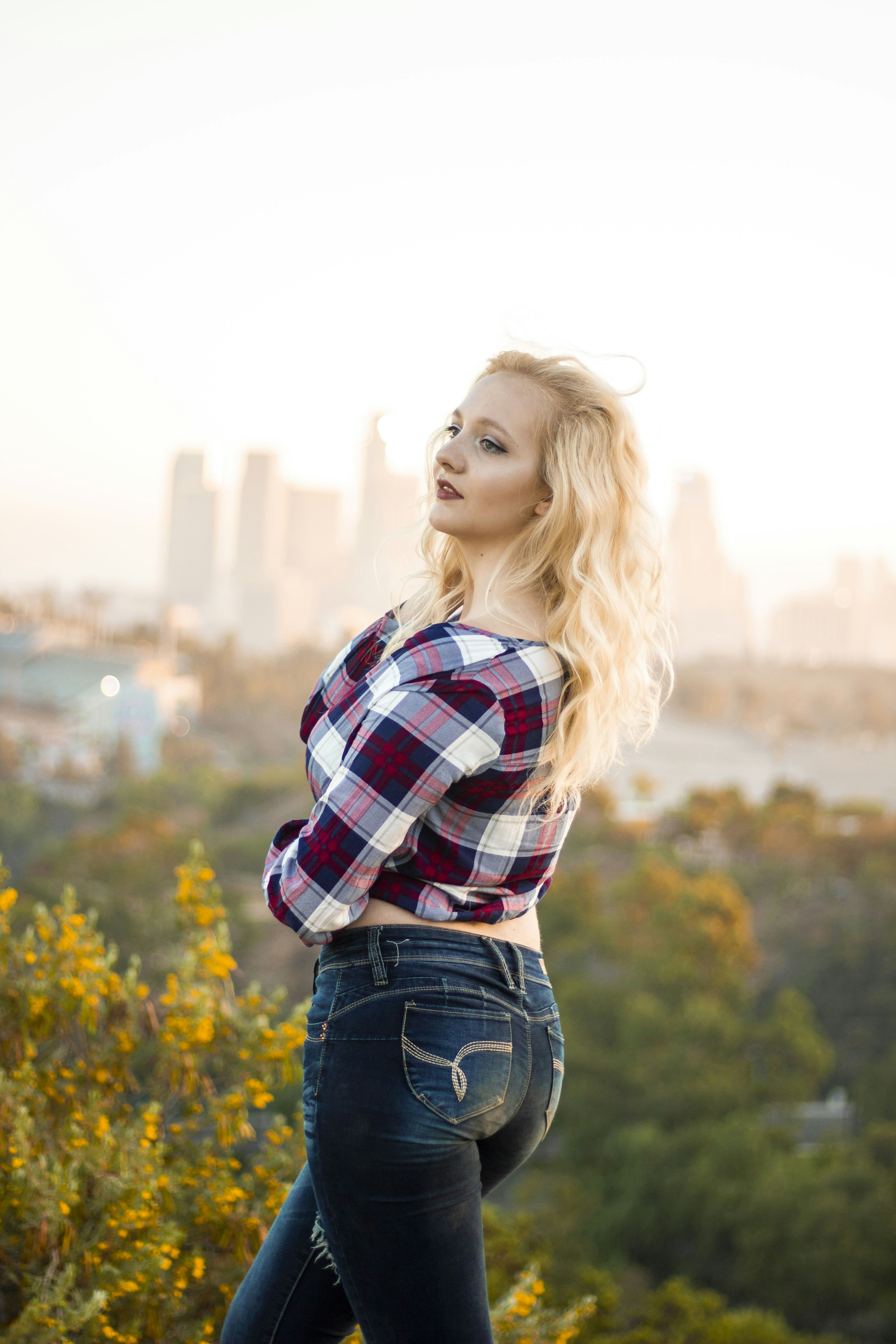 Blonde woman in a plaid shirt poses against a backdrop of a city skyline, showcasing the contrast between nature and urban life.