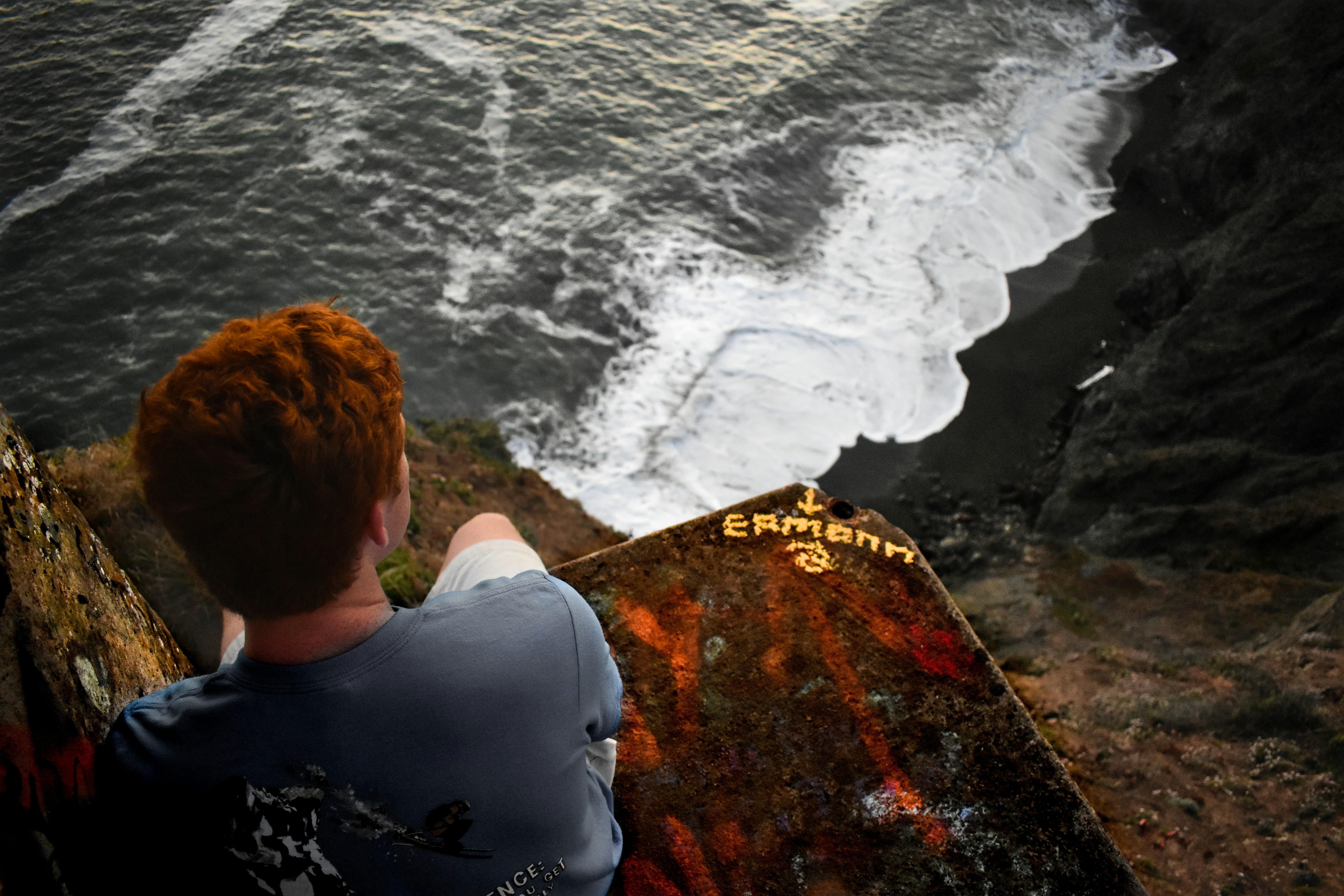 A young man sits on a rocky ledge overlooking crashing waves below, with vibrant graffiti nearby. The scene captures a moment of contemplation against a dramatic coastal backdrop.