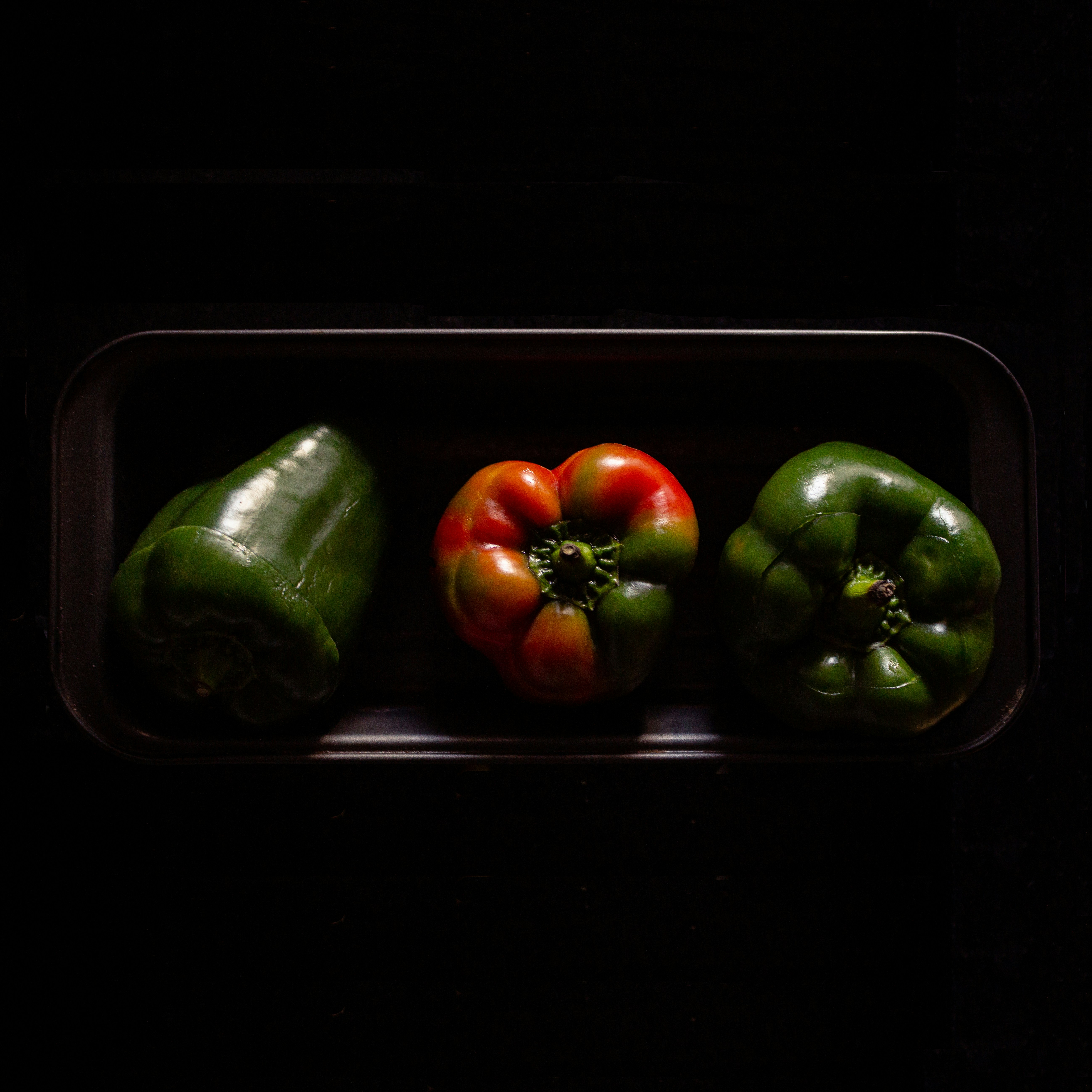 Three bell peppers arranged in a sleek tray: a green pepper, a red and yellow pepper, and another green pepper, all against a dark background.