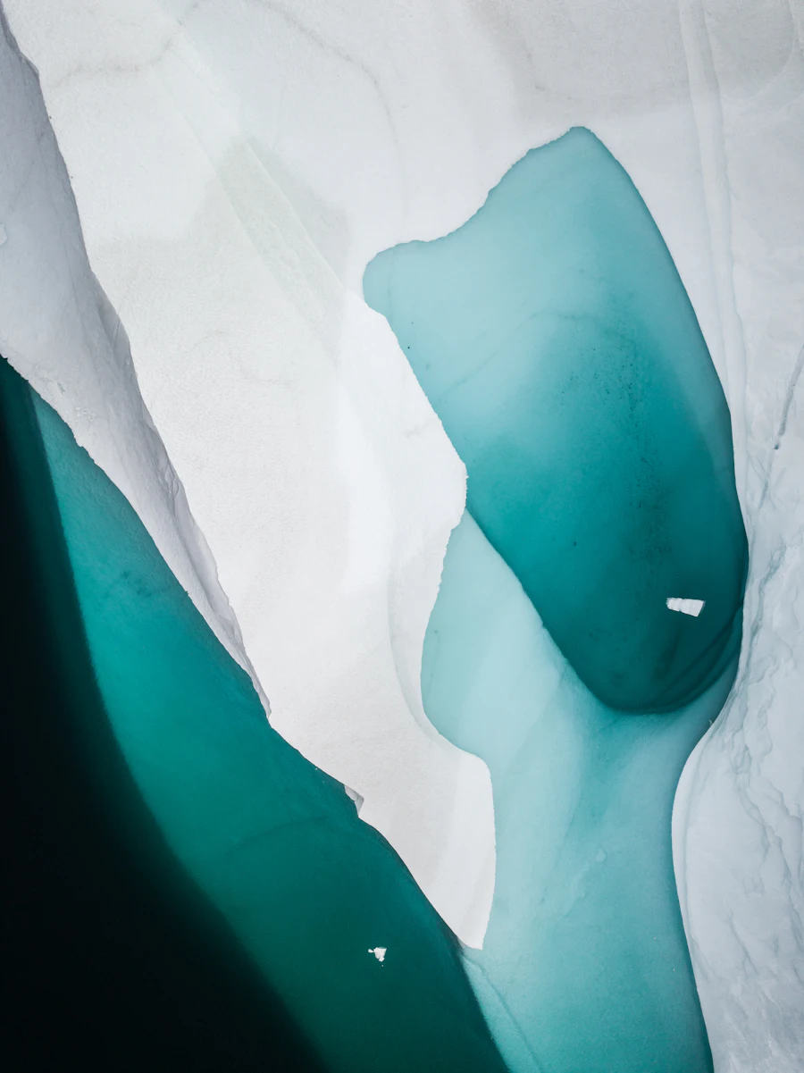 Abstract aerial view of white ice fields framing vivid turquoise meltwater pools and dark crevasses.