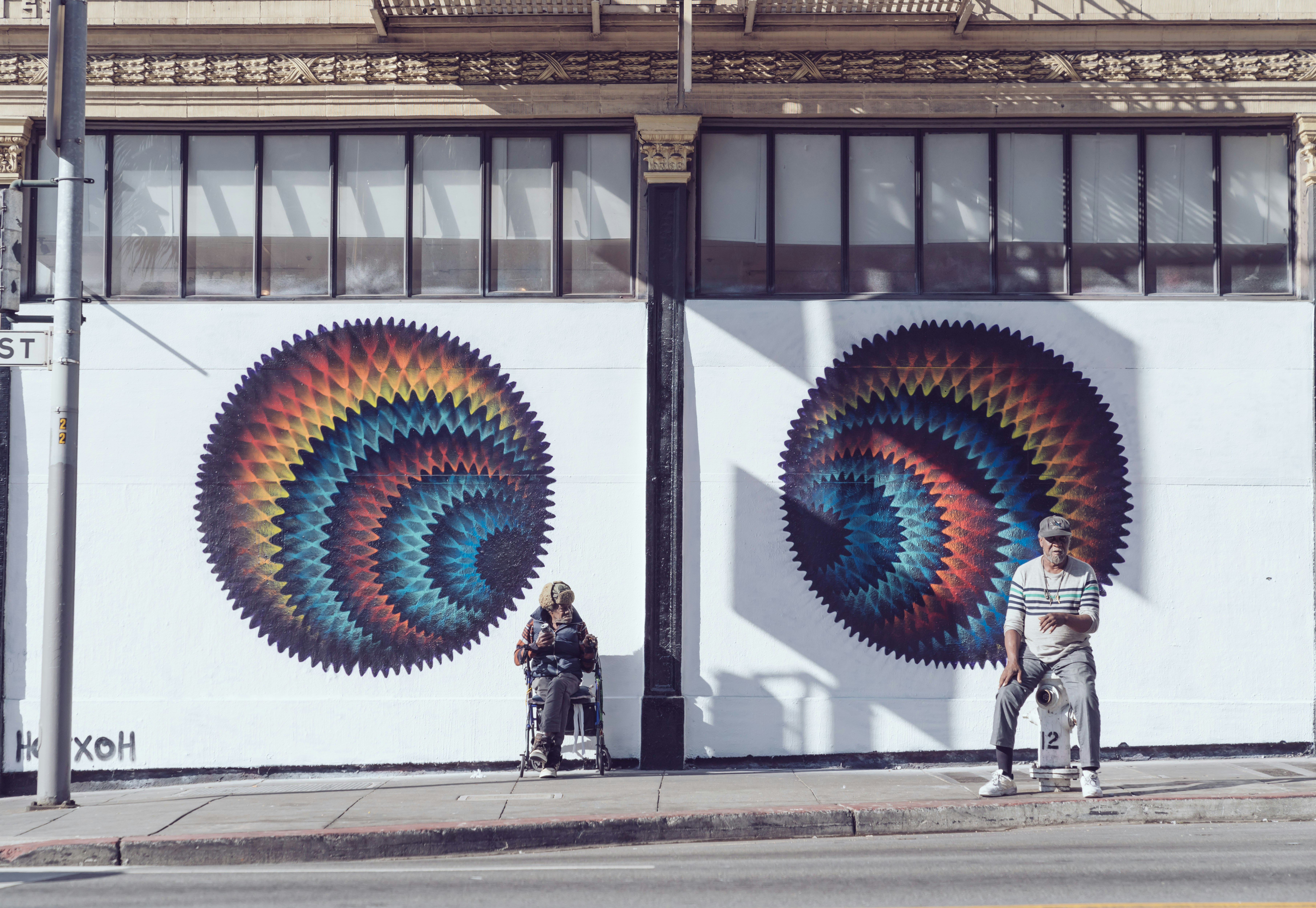 Vibrant mural featuring swirling patterns on a wall, flanked by two seated figures. The artwork draws the eye with its dynamic colors and shapes.