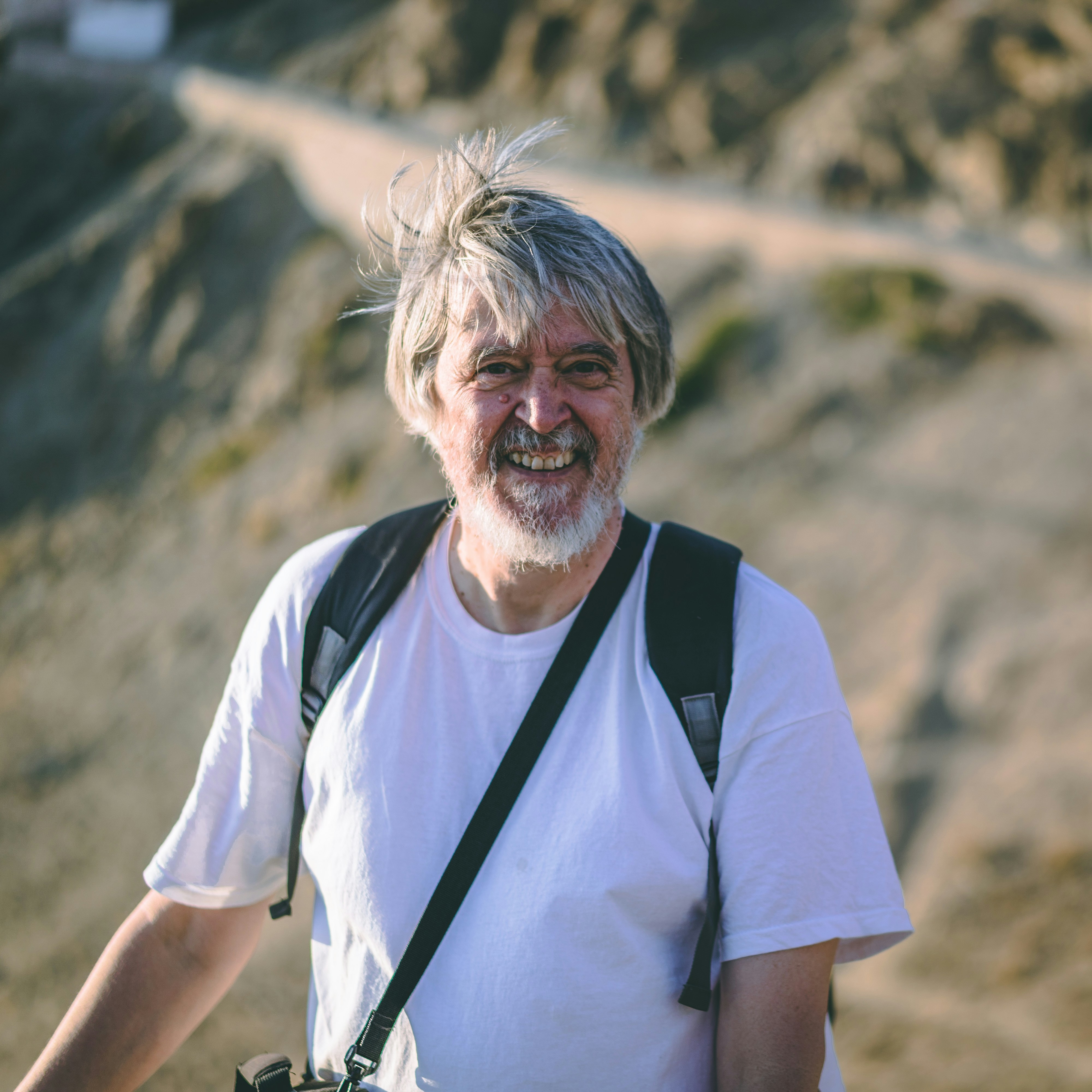 Smiling man with tousled hair stands on a ridge, basking in the sunlight with a scenic backdrop of hills and paths.