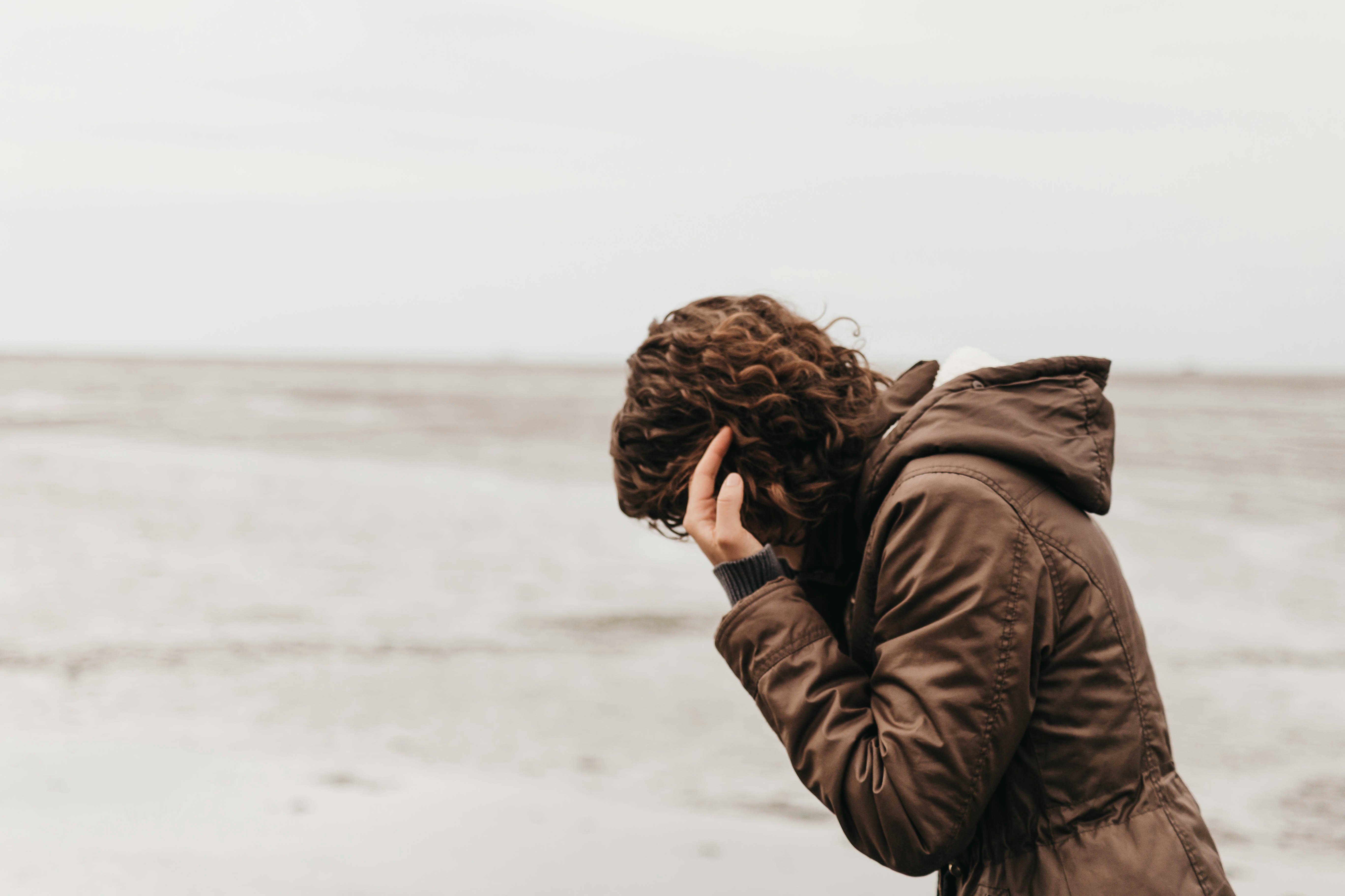 A woman in a brown jacket stands by a vast, muted shoreline, her hand resting on her head in contemplation. The subdued colors evoke a sense of introspection.