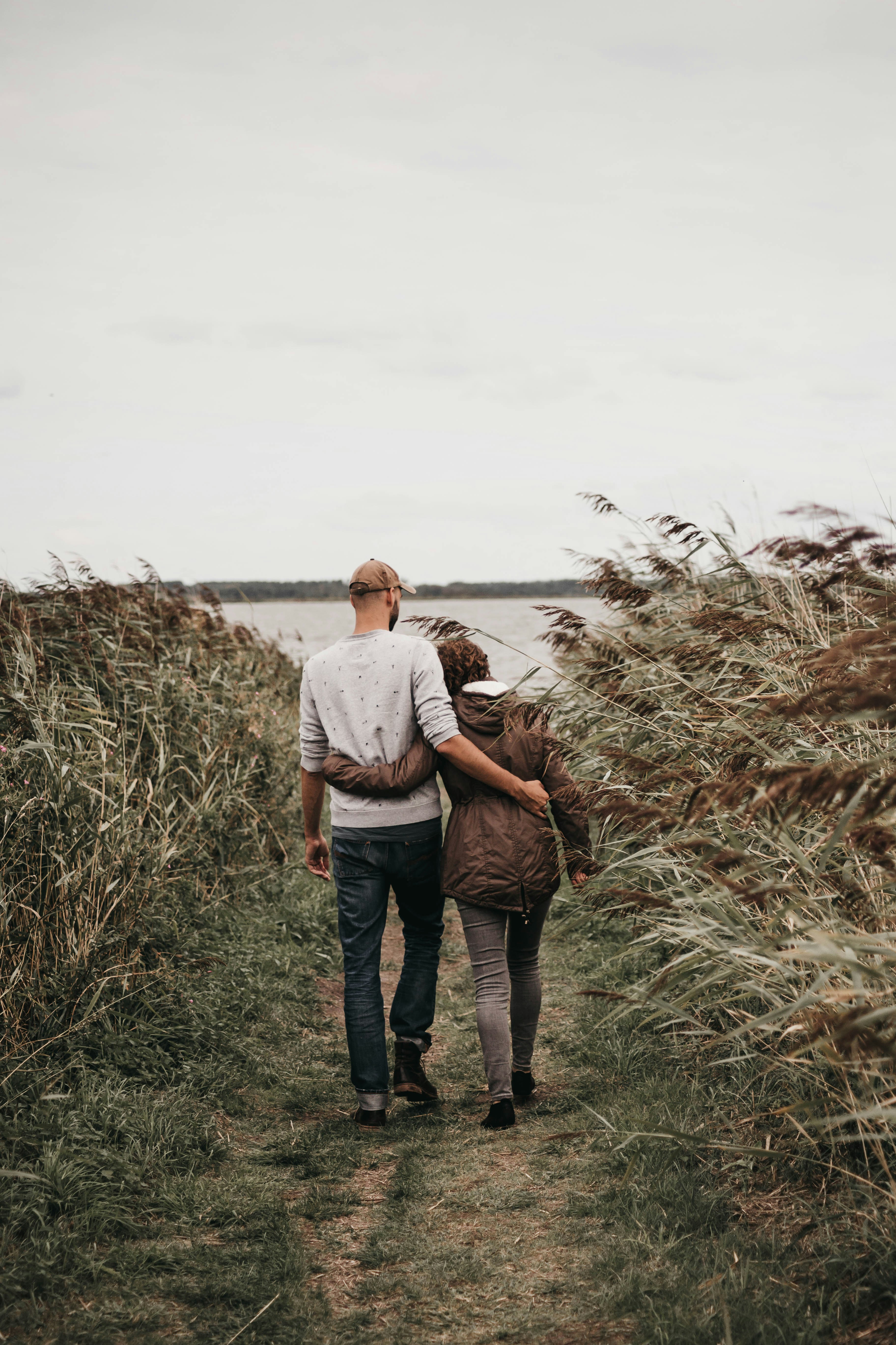 Couple walking hand-in-hand along a grassy path bordered by tall reeds, leading to a serene body of water in the distance.