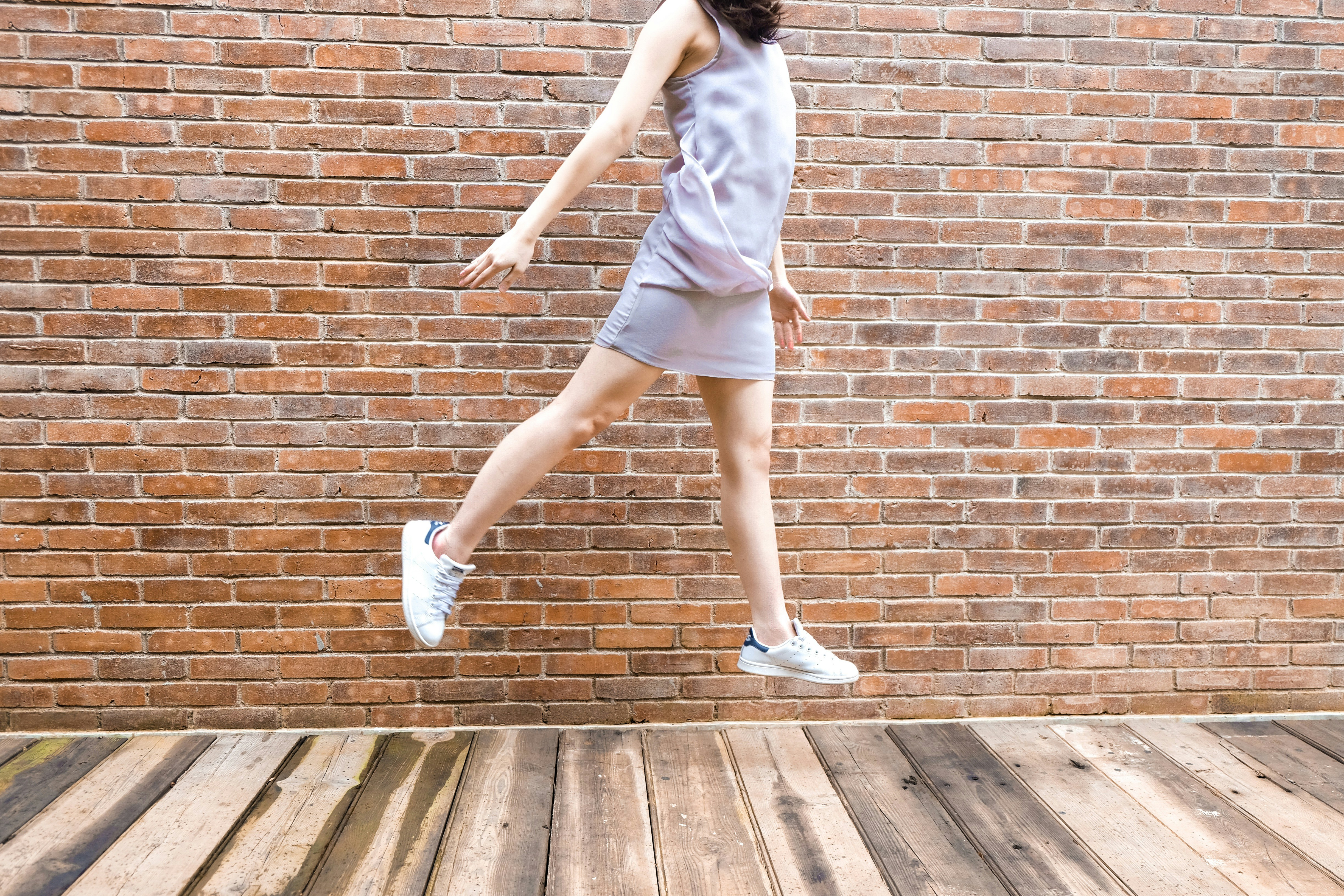 Person jumping in mid-air next to a brick wall, wearing a light dress and sneakers on a wooden floor.
