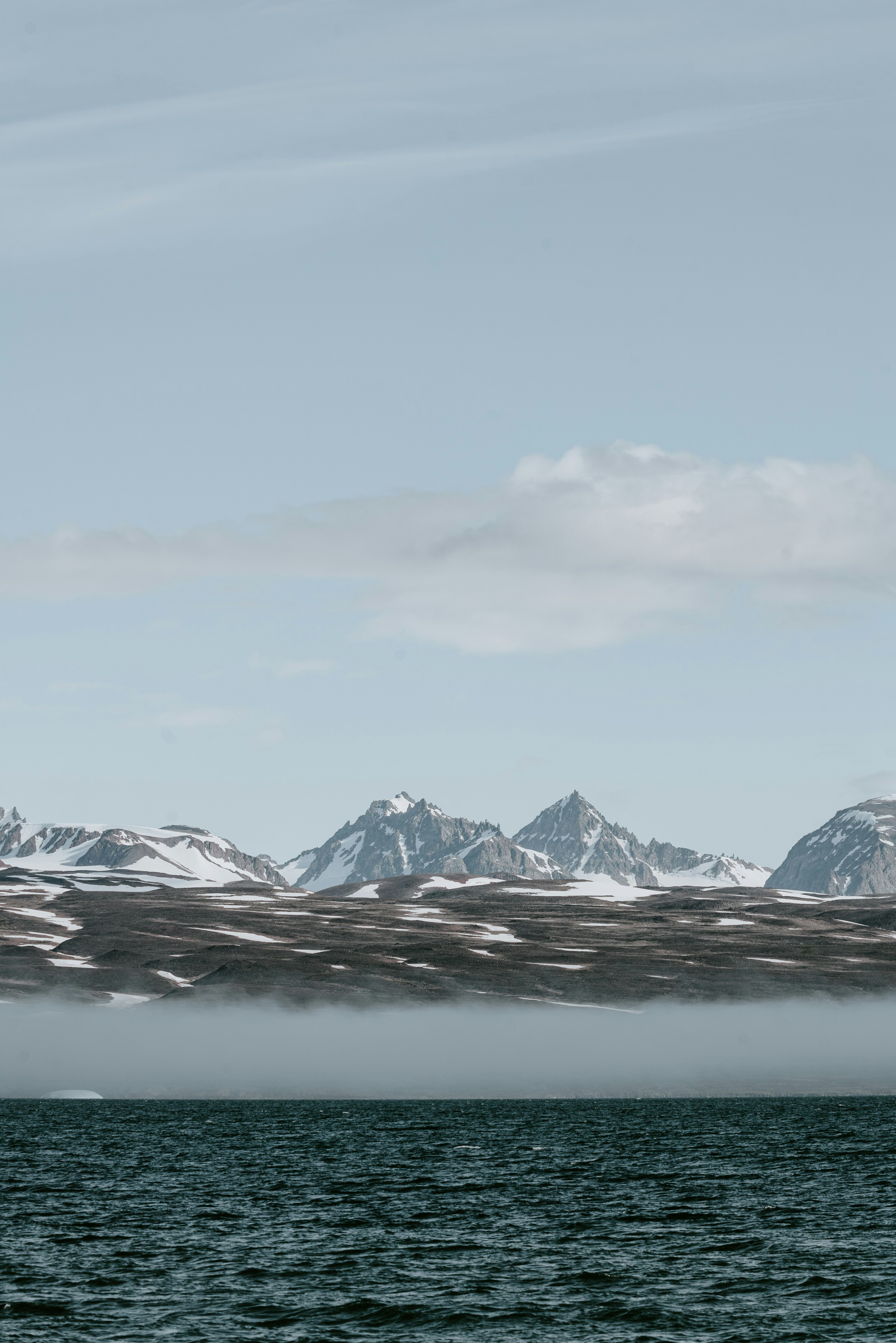 Snow-capped mountains rise majestically above a tranquil sea, shrouded in mist under a pale sky.