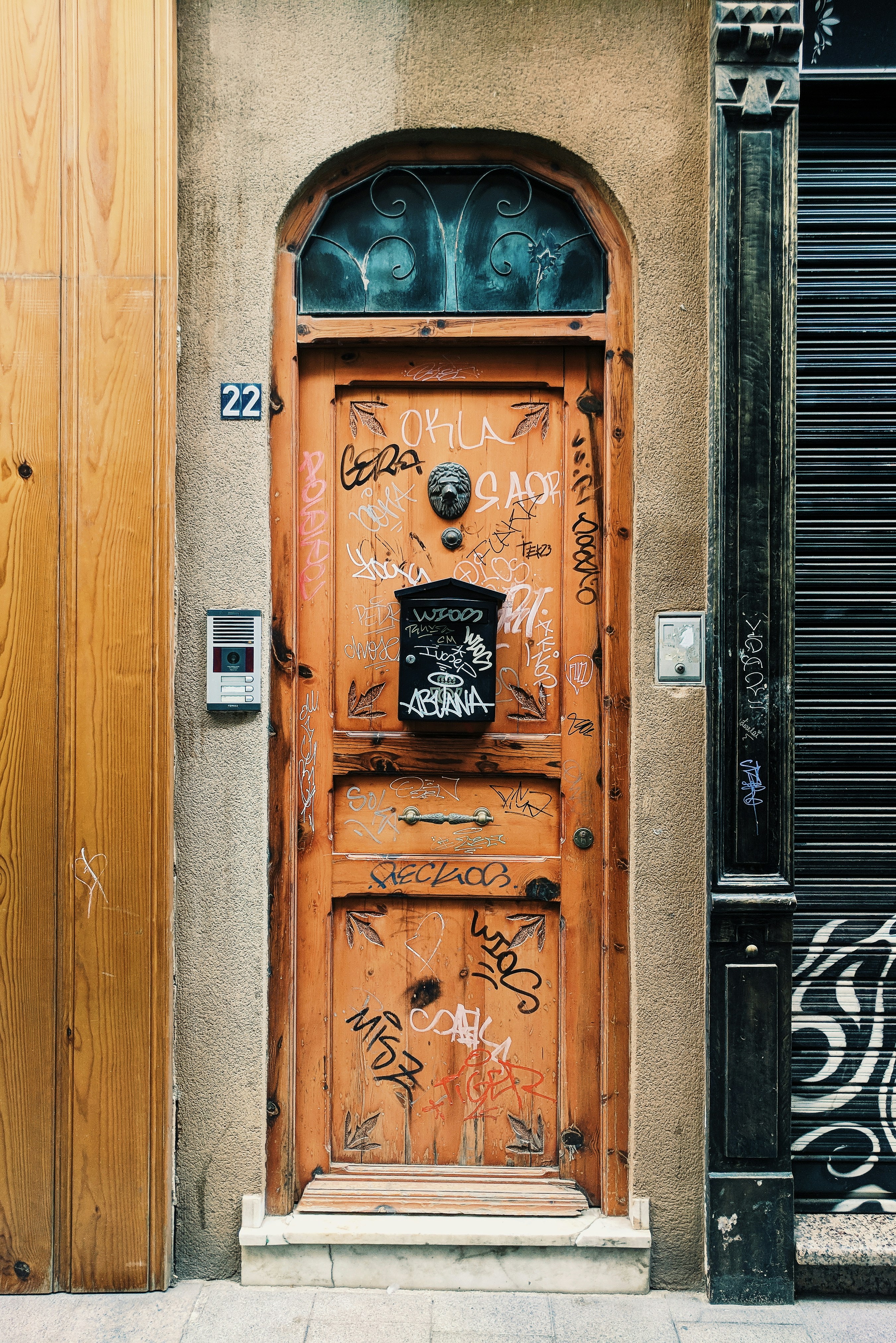Weathered orange door covered in graffiti sits between a wooden panel on the left and a dark metal shutter on the right. A small intercom panel and a graffiti-tagged frame highlight the gritty urban setting.