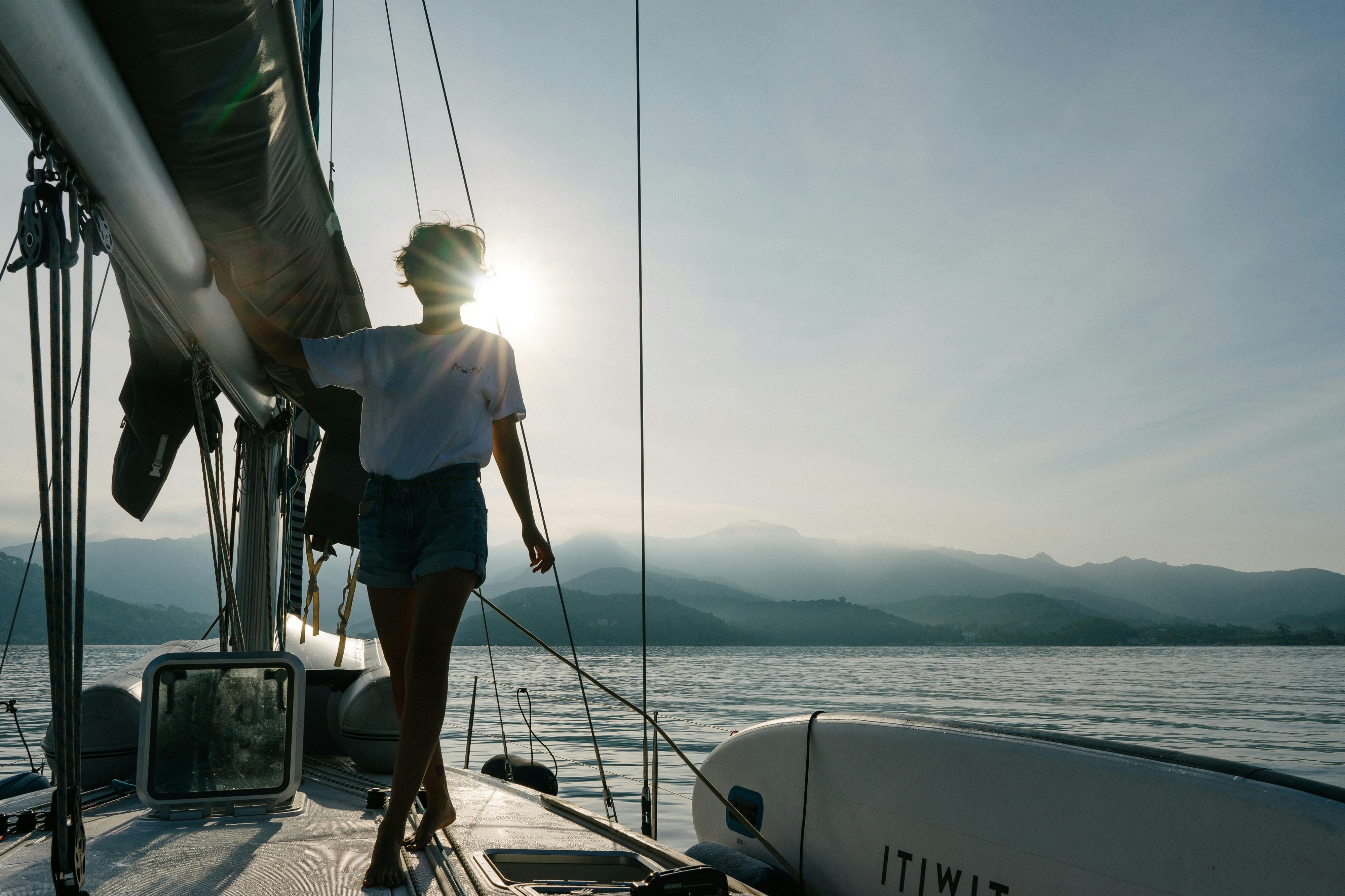 Silhouetted woman stands on a sailboat with the sun setting behind, casting a serene glow over distant mountains and calm waters.