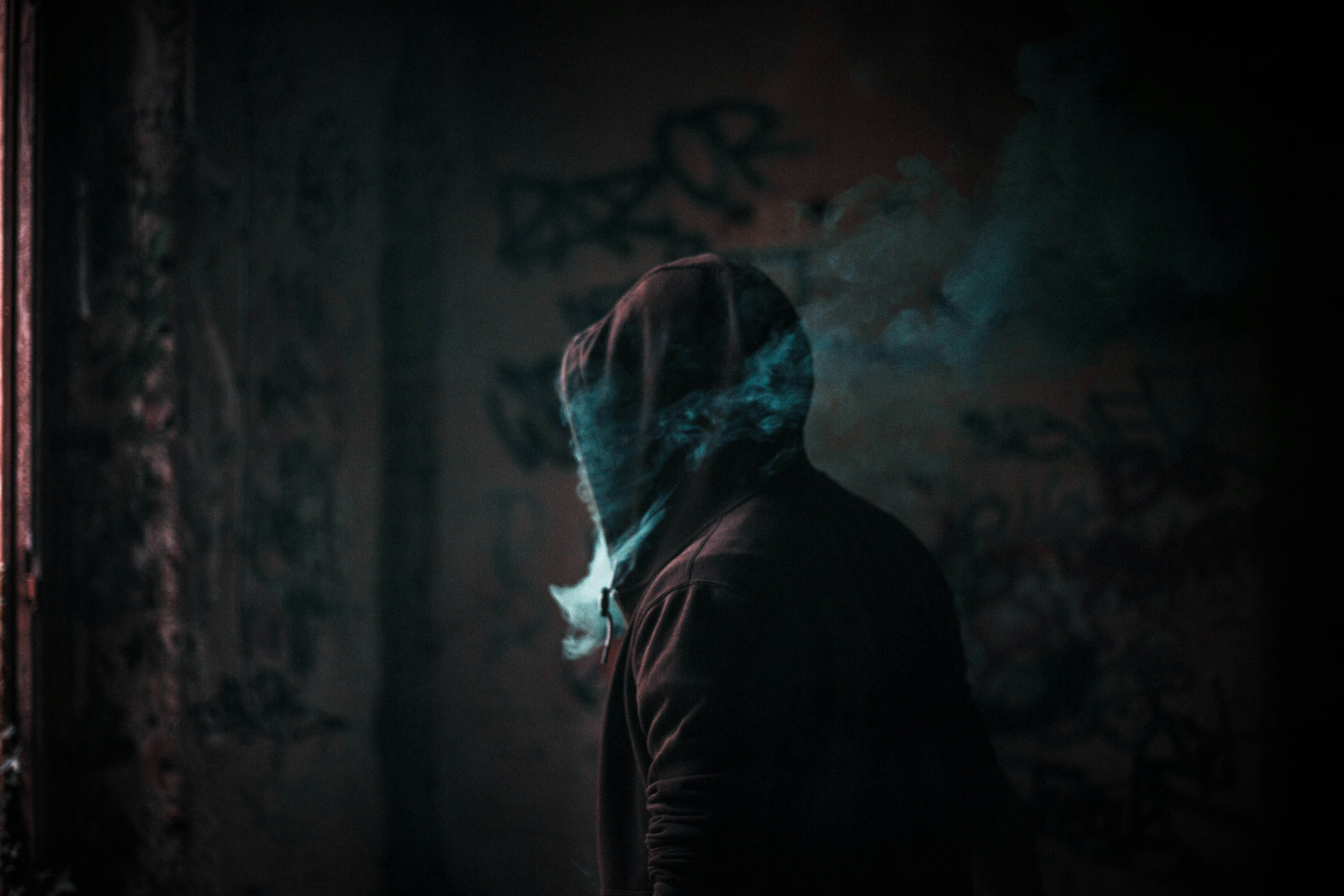 A hooded figure stands in a dimly lit, graffiti-covered space, exhaling a plume of smoke. The atmosphere is thick with mystery and urban decay.