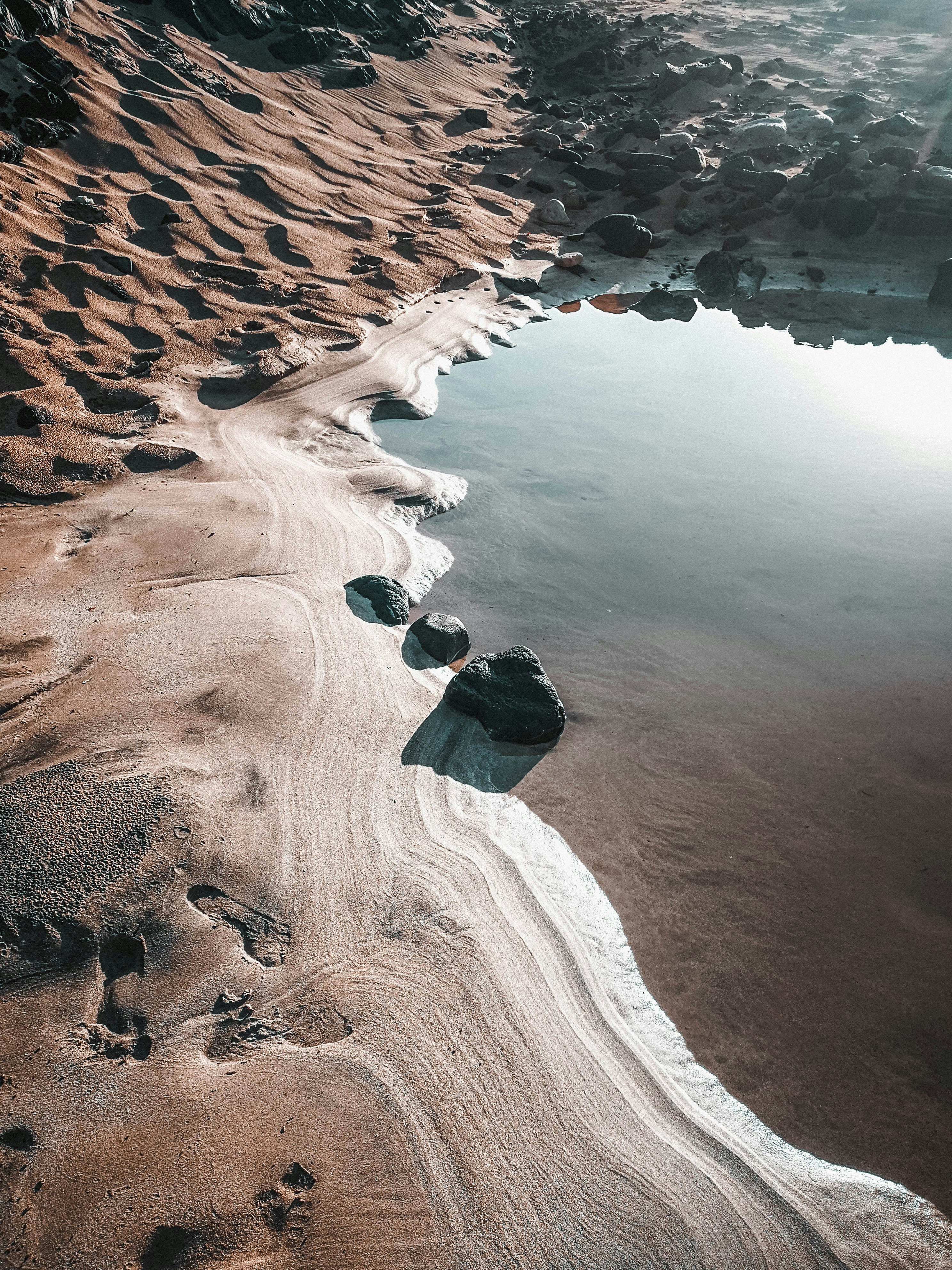 Gentle curves of sand and water reveal a tranquil shoreline, accentuated by scattered stones. The reflective surface mirrors the serene environment.