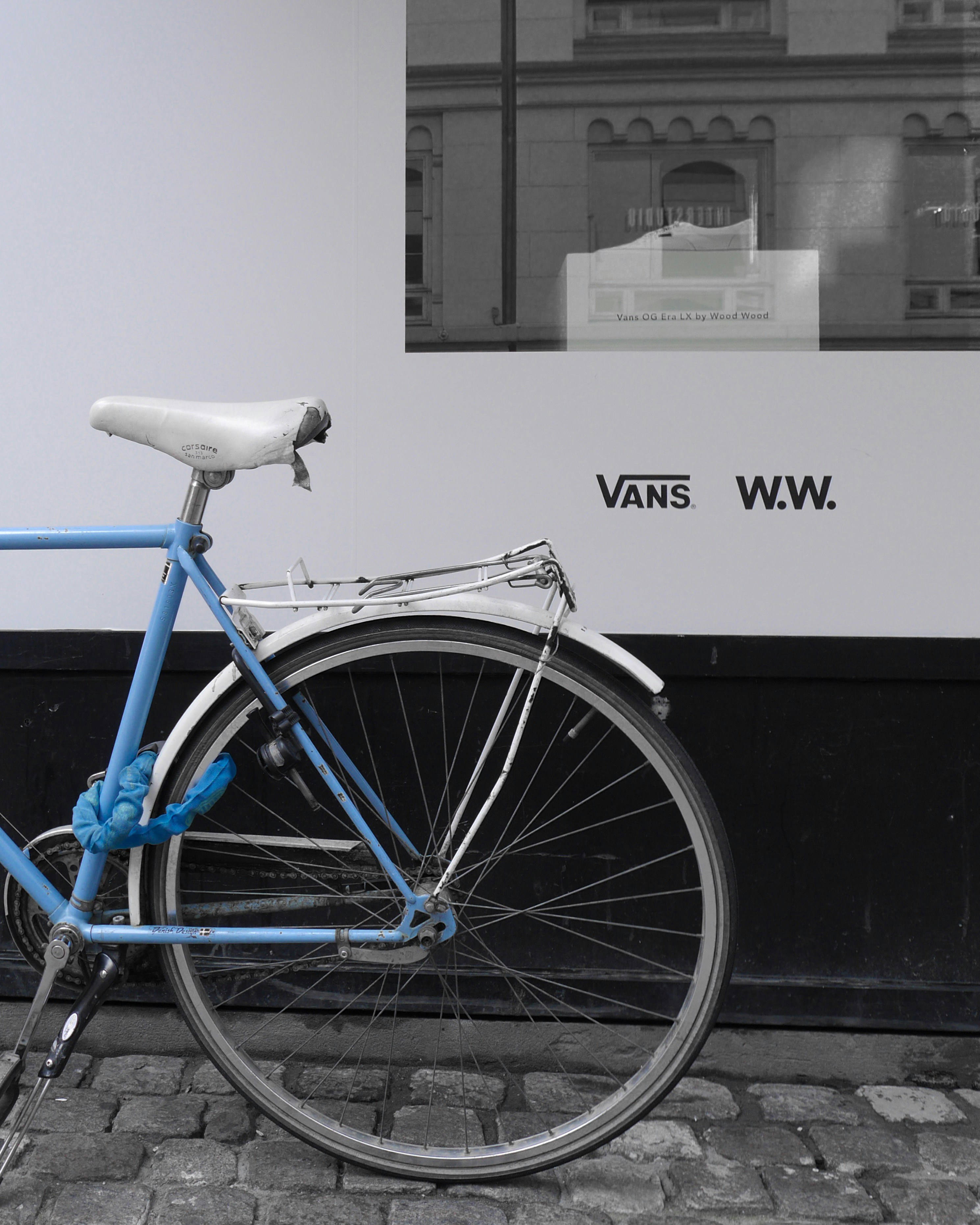 A blue bicycle secured with a blue lock stands against a minimalist white wall featuring logo elements. The composition highlights urban life and design.