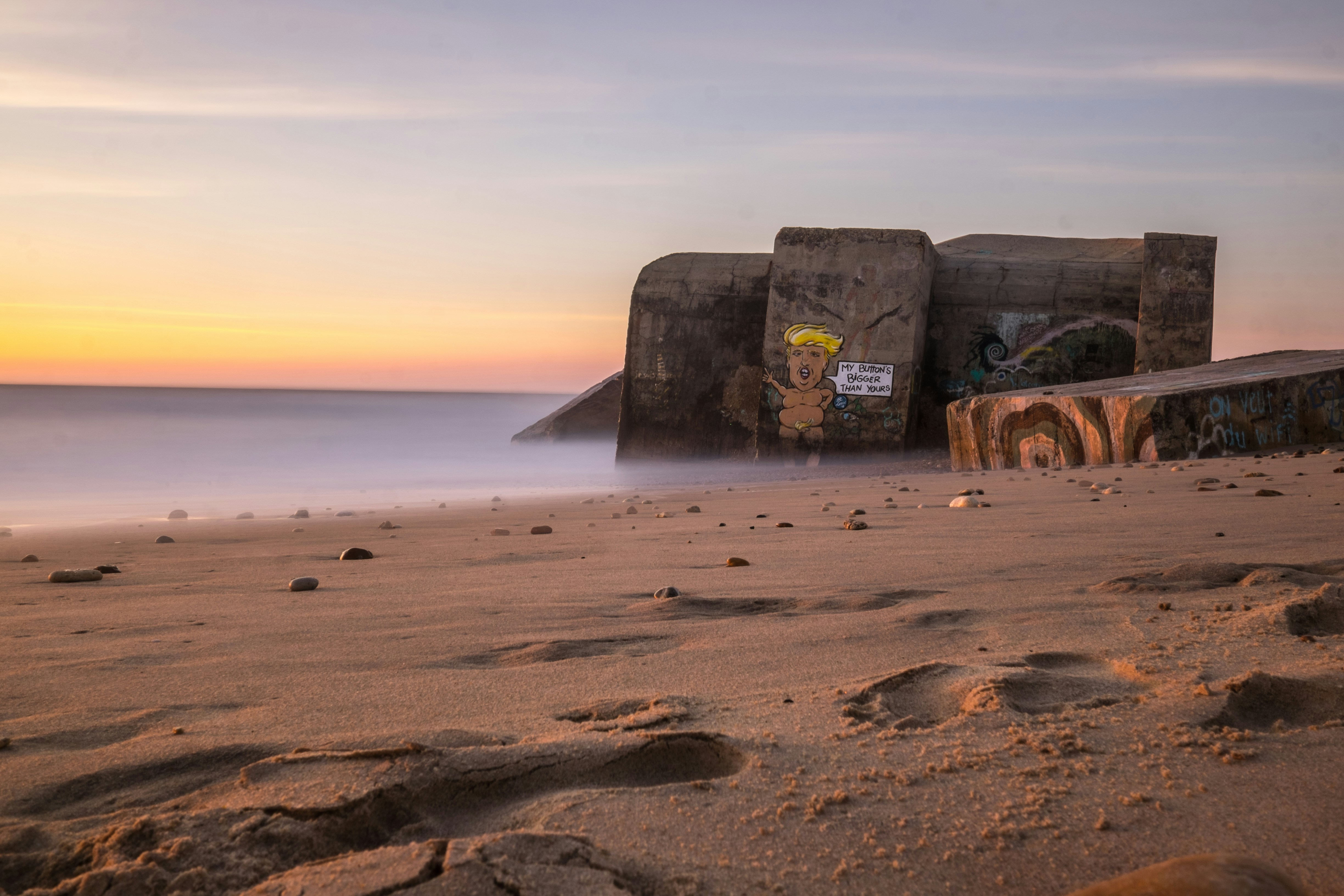 Weathered concrete blocks adorned with graffiti stand against a serene beach backdrop at sunset, capturing the intersection of art and nature.