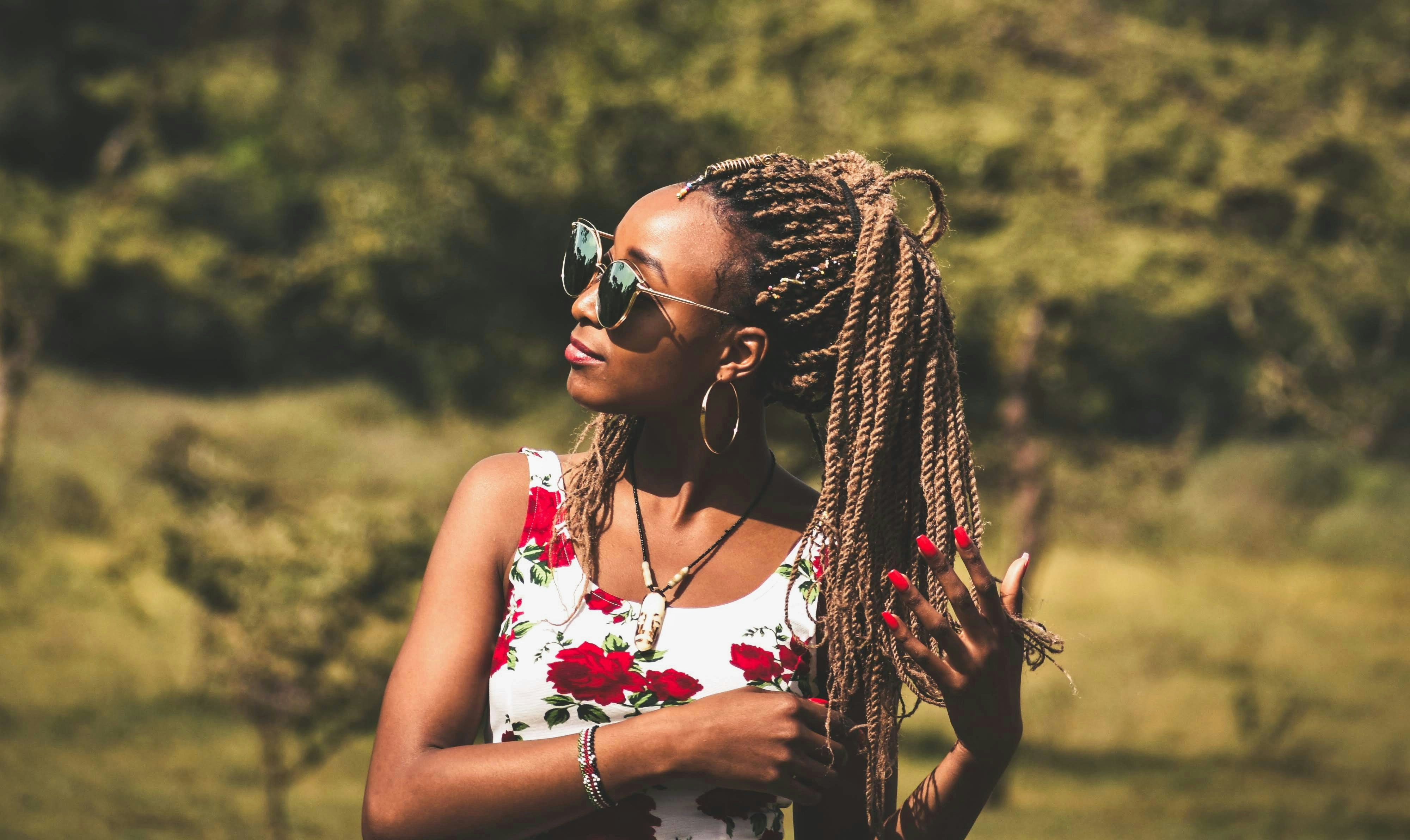 Woman in floral dress with braided hair stands in sunlight against a lush green backdrop.
