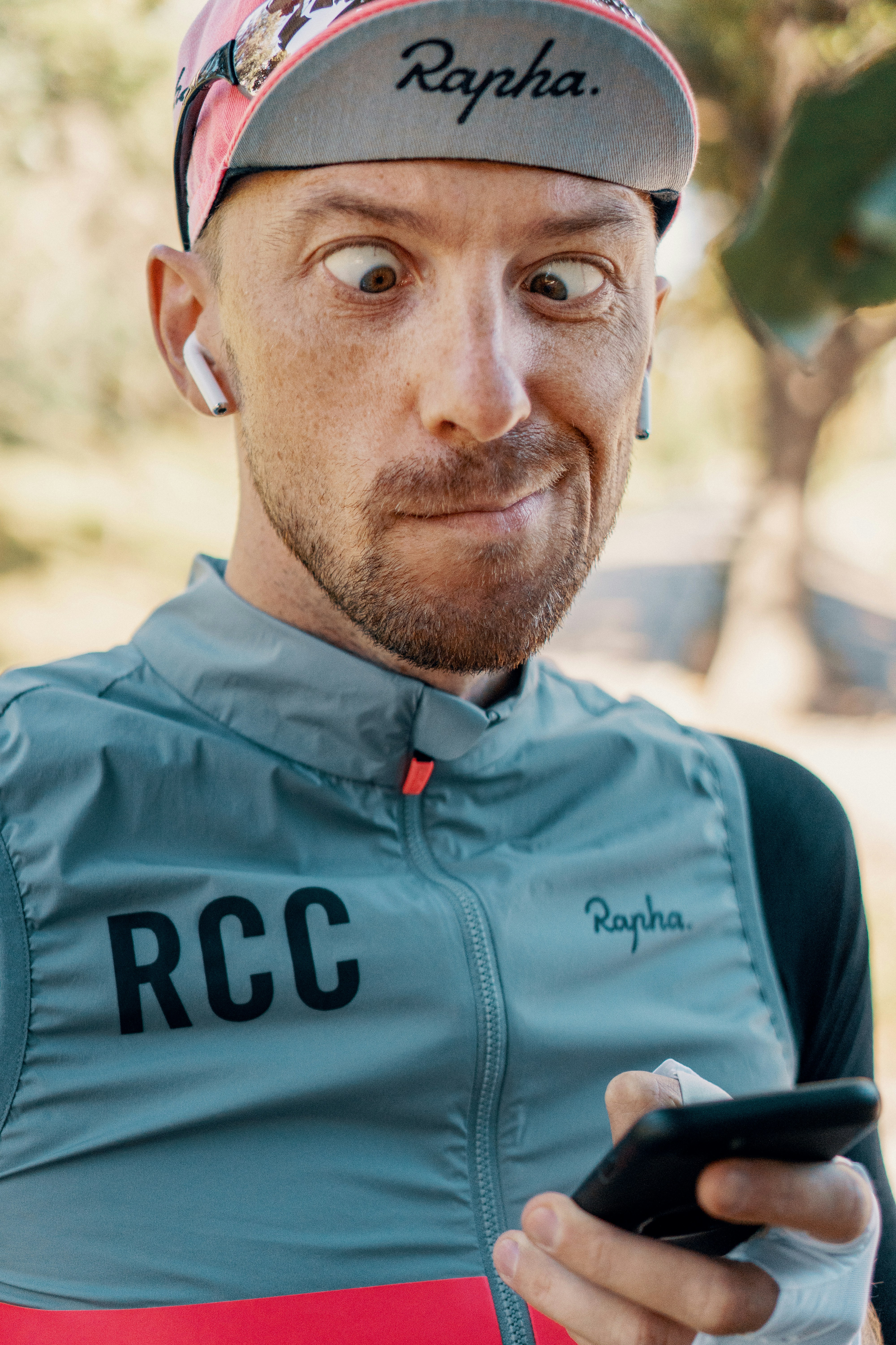 Cyclist with a playful expression checks his phone while wearing a Rapha cycling outfit and cap. The background features soft natural greenery.