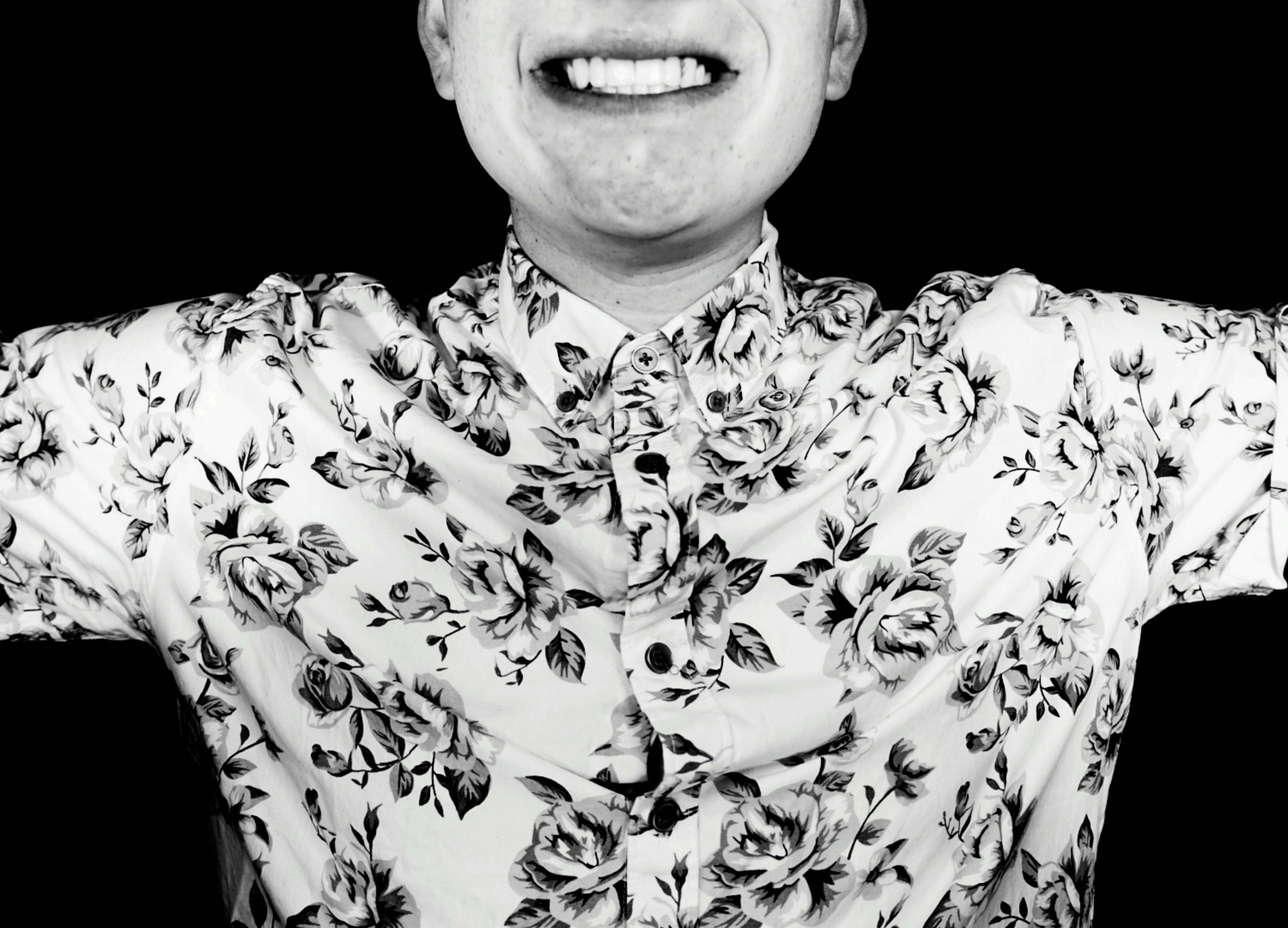 Person in a floral shirt smiling widely with arms outstretched against a black background.